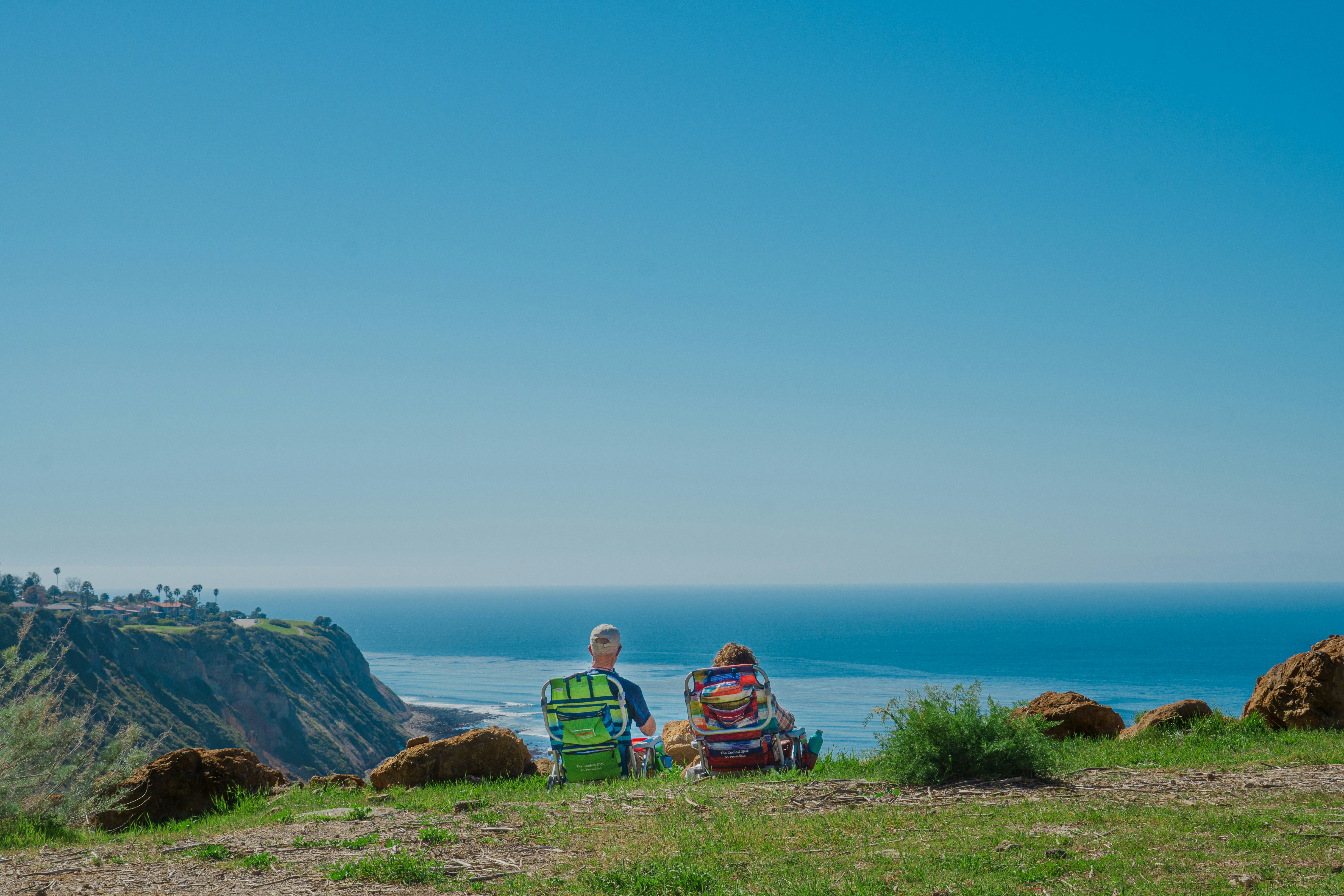 Two people relax on a grassy cliff overlooking the deep blue ocean under a clear sky.