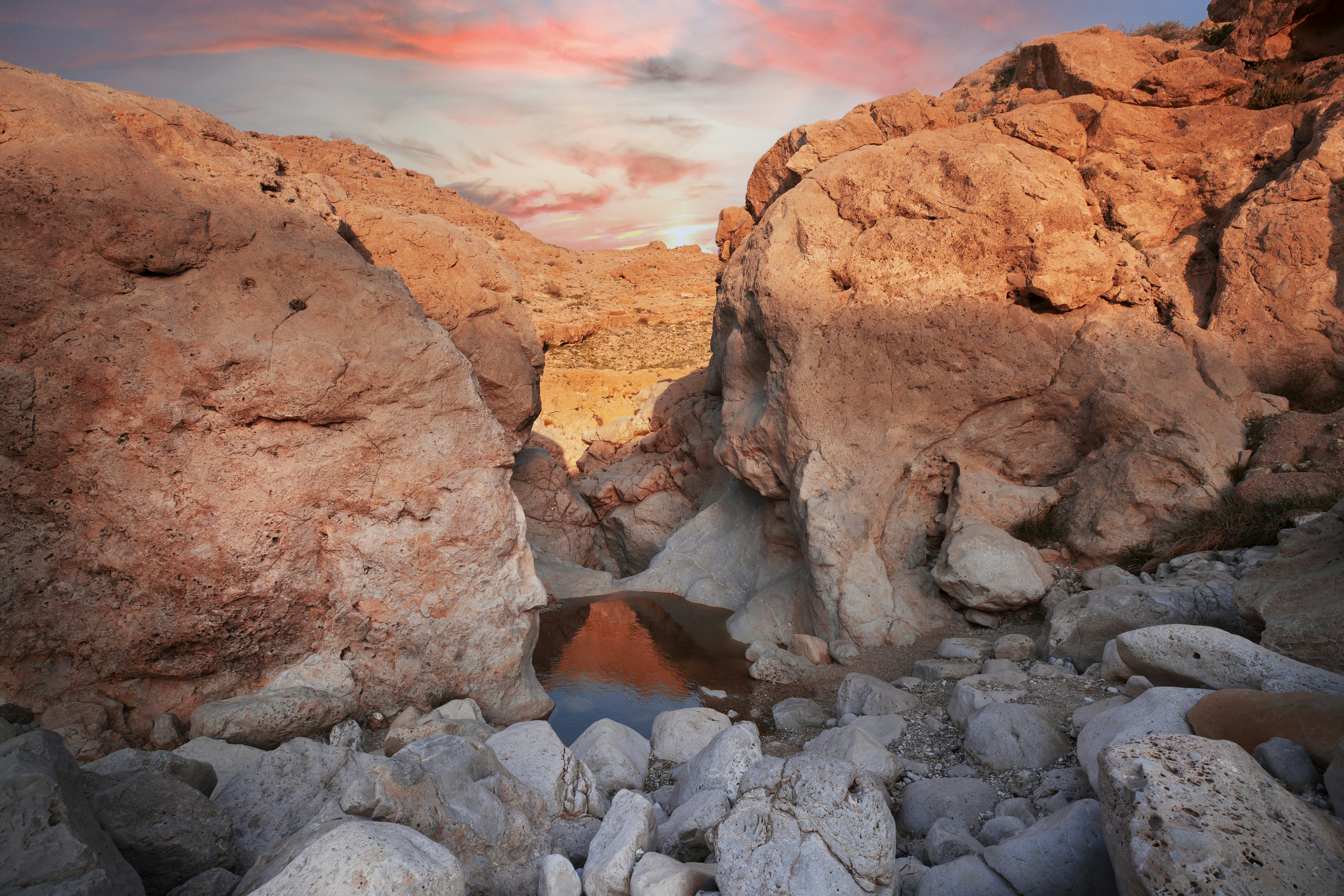 Rocky canyon illuminated by warm sunset hues with a tranquil water pool reflecting the sky.
