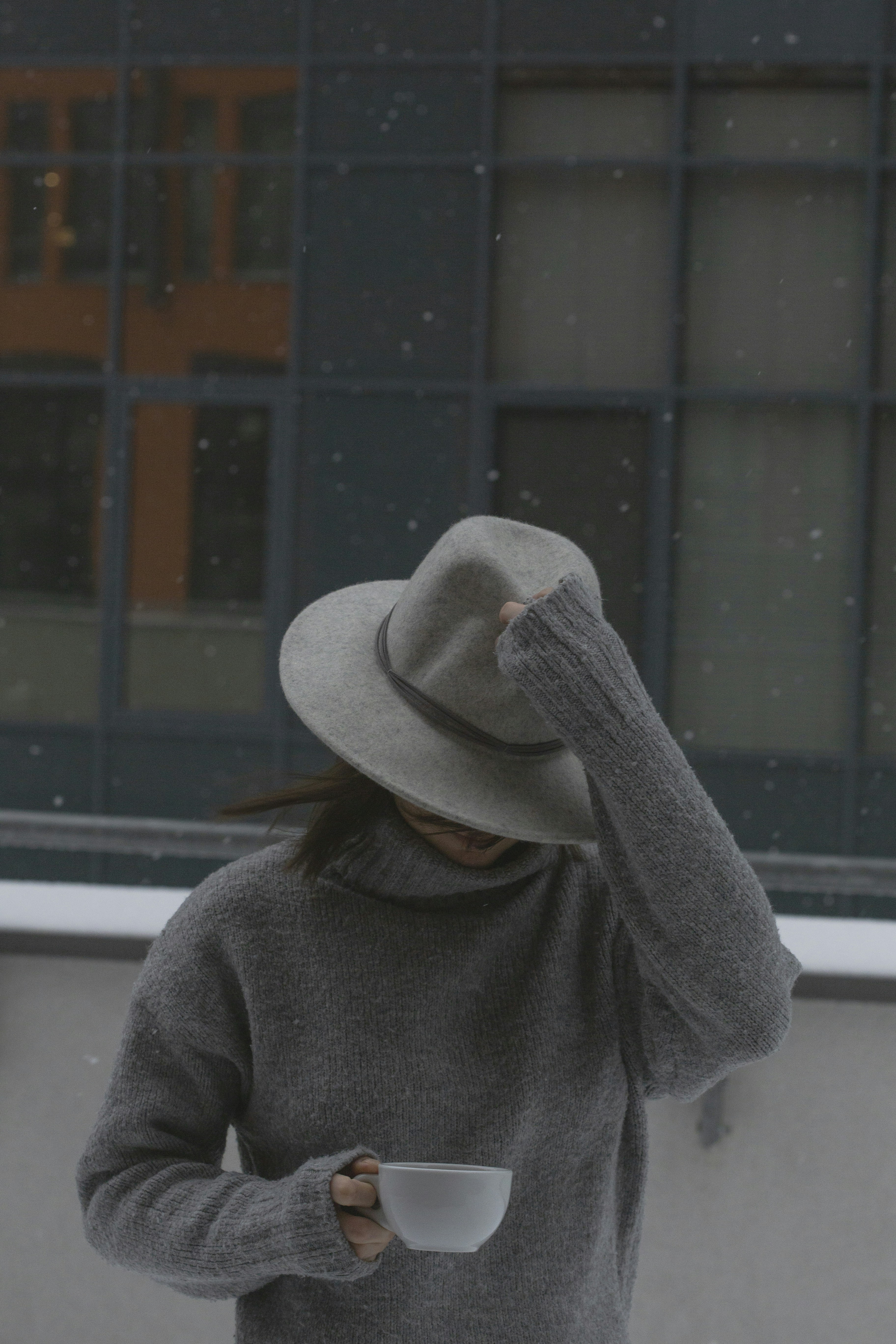 Woman in a gray sweater and hat, holding a white cup, amidst falling snowflakes on a rooftop. Urban backdrop enhances the winter ambiance.