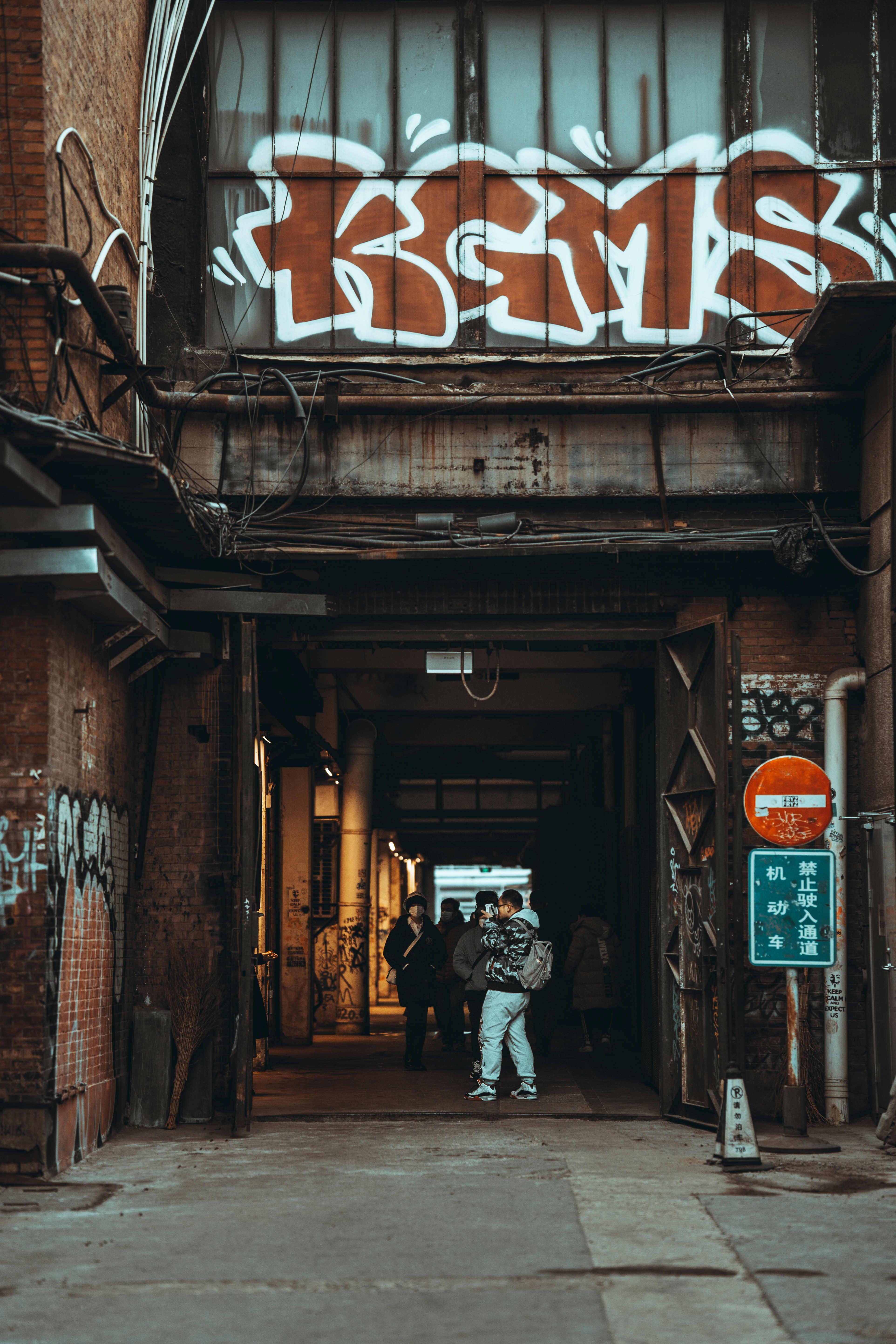 Graffiti adorns the walls of a dimly lit urban passageway, where figures gather in conversation under the glow of distant lights.