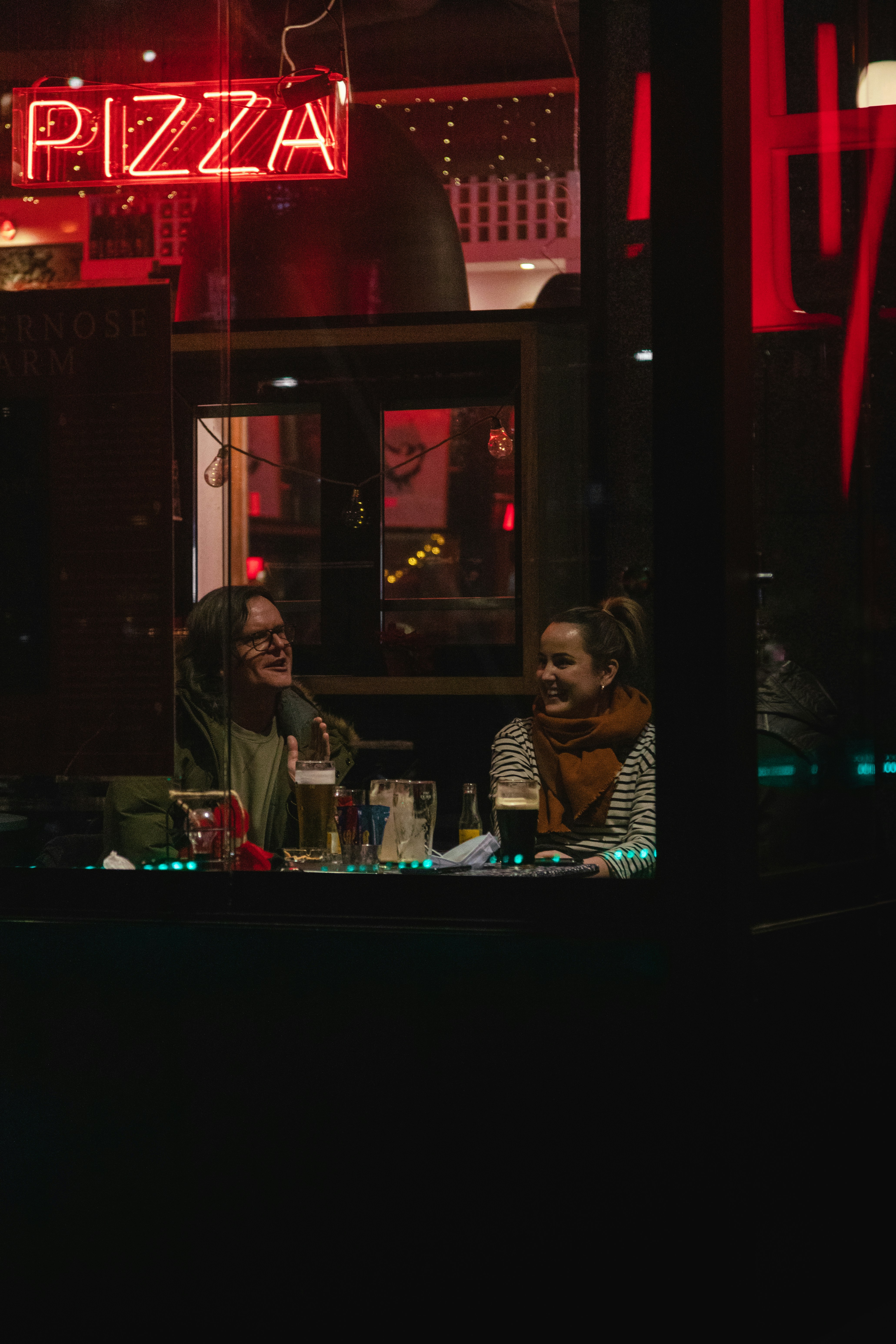 Two friends enjoying pizza and drinks in a warmly lit restaurant, highlighted by a glowing neon sign. The ambiance captures a cozy night out.