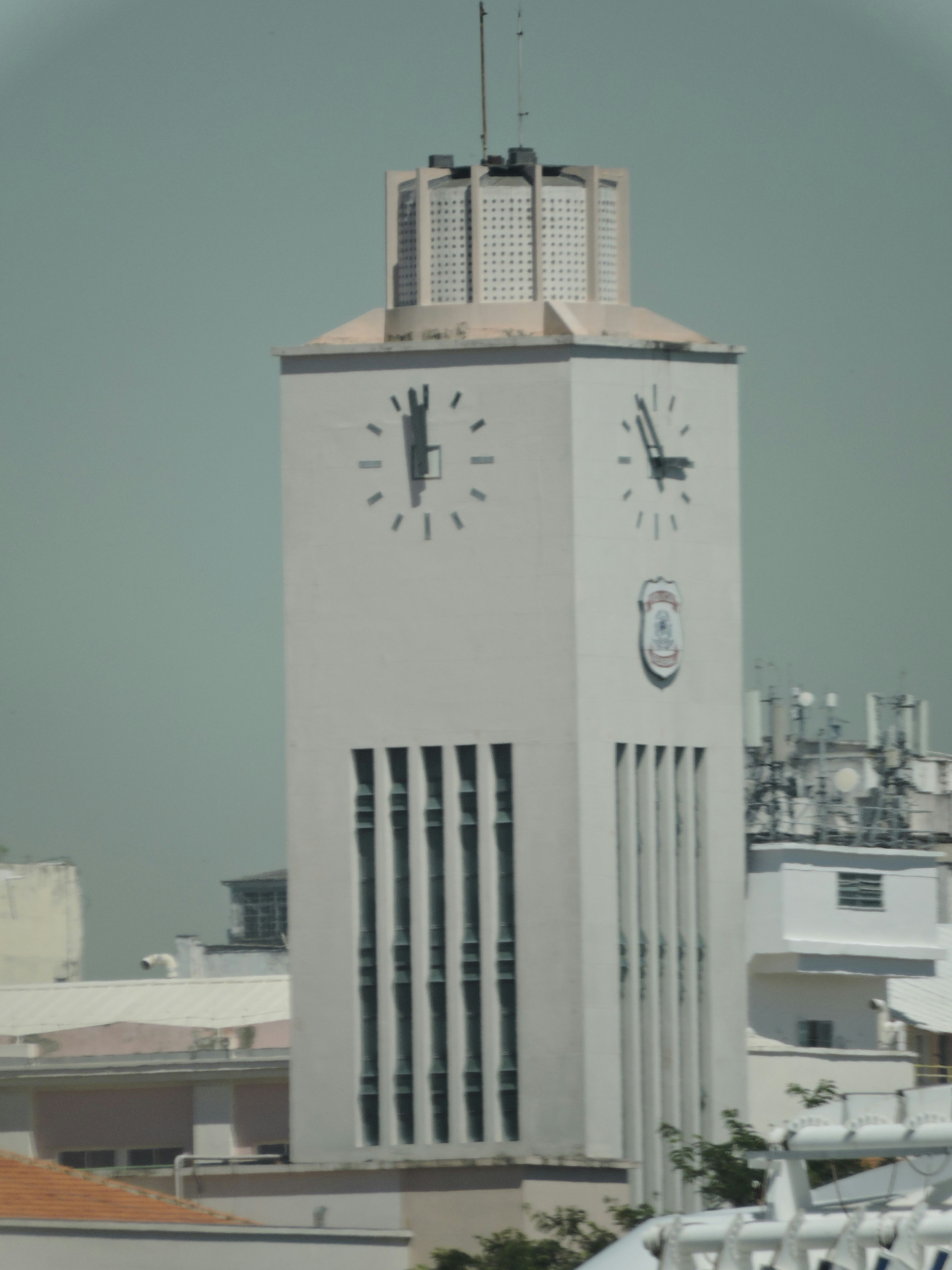 Photograph showing a tall clocktower with twin clocks against a muted teal sky. The tower’s white façade and surrounding rooftops anchor the urban scene.