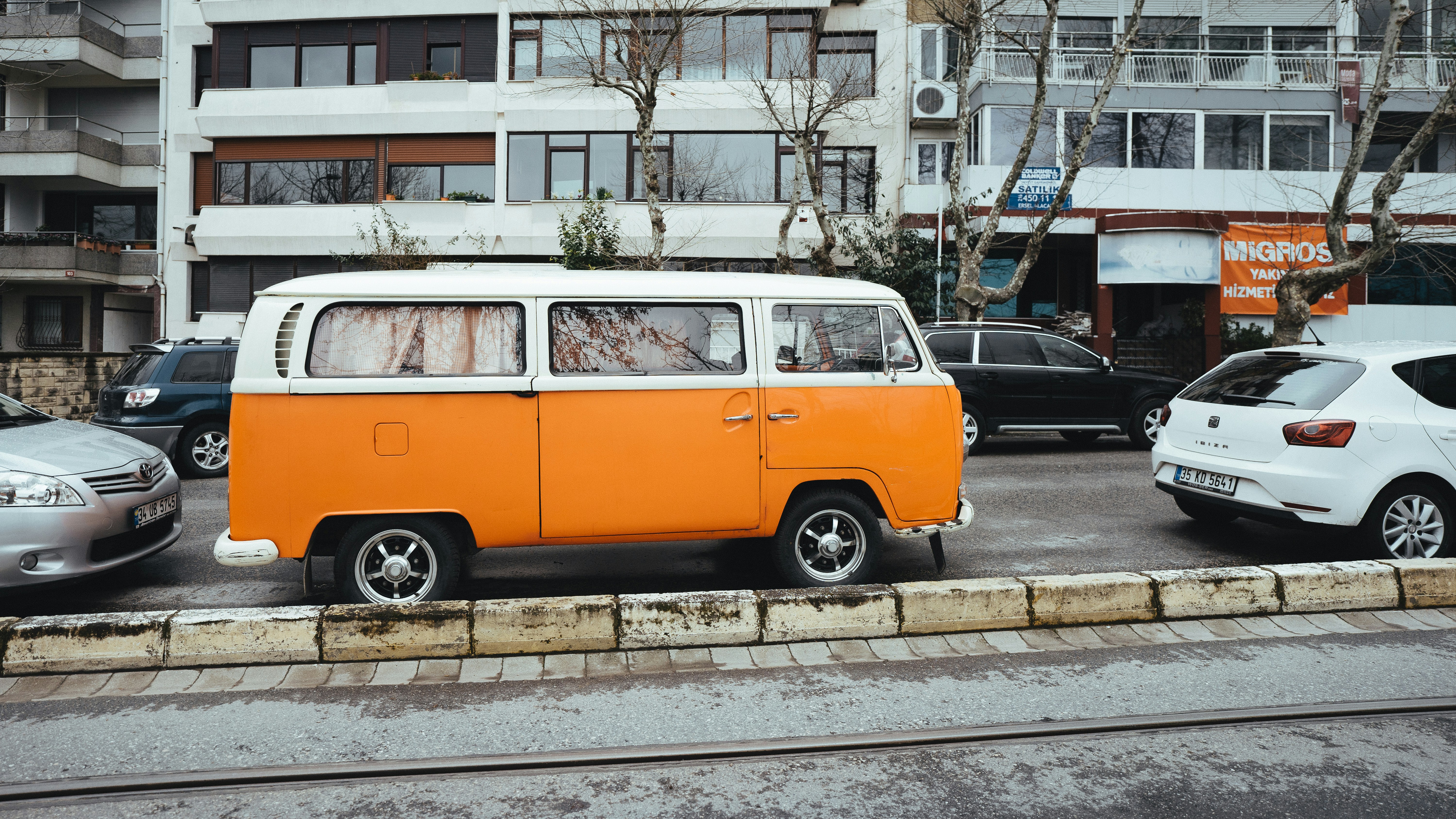 A classic orange and white VW van parked alongside contemporary vehicles on a bustling street. The scene captures a blend of nostalgia and urban life.