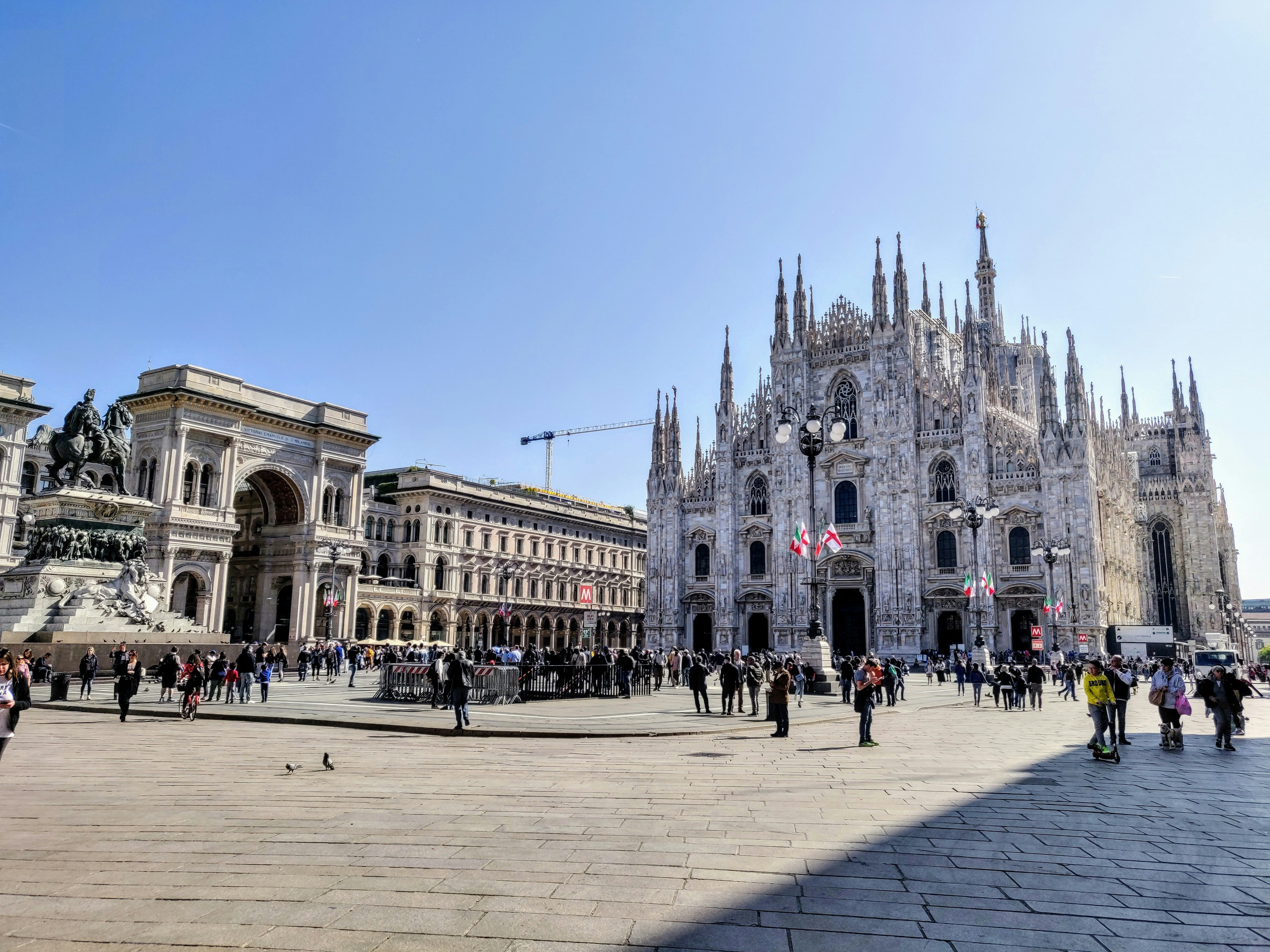 Gothic cathedral towering over bustling square, surrounded by historic buildings and lively crowds. Flags flutter in the breeze.