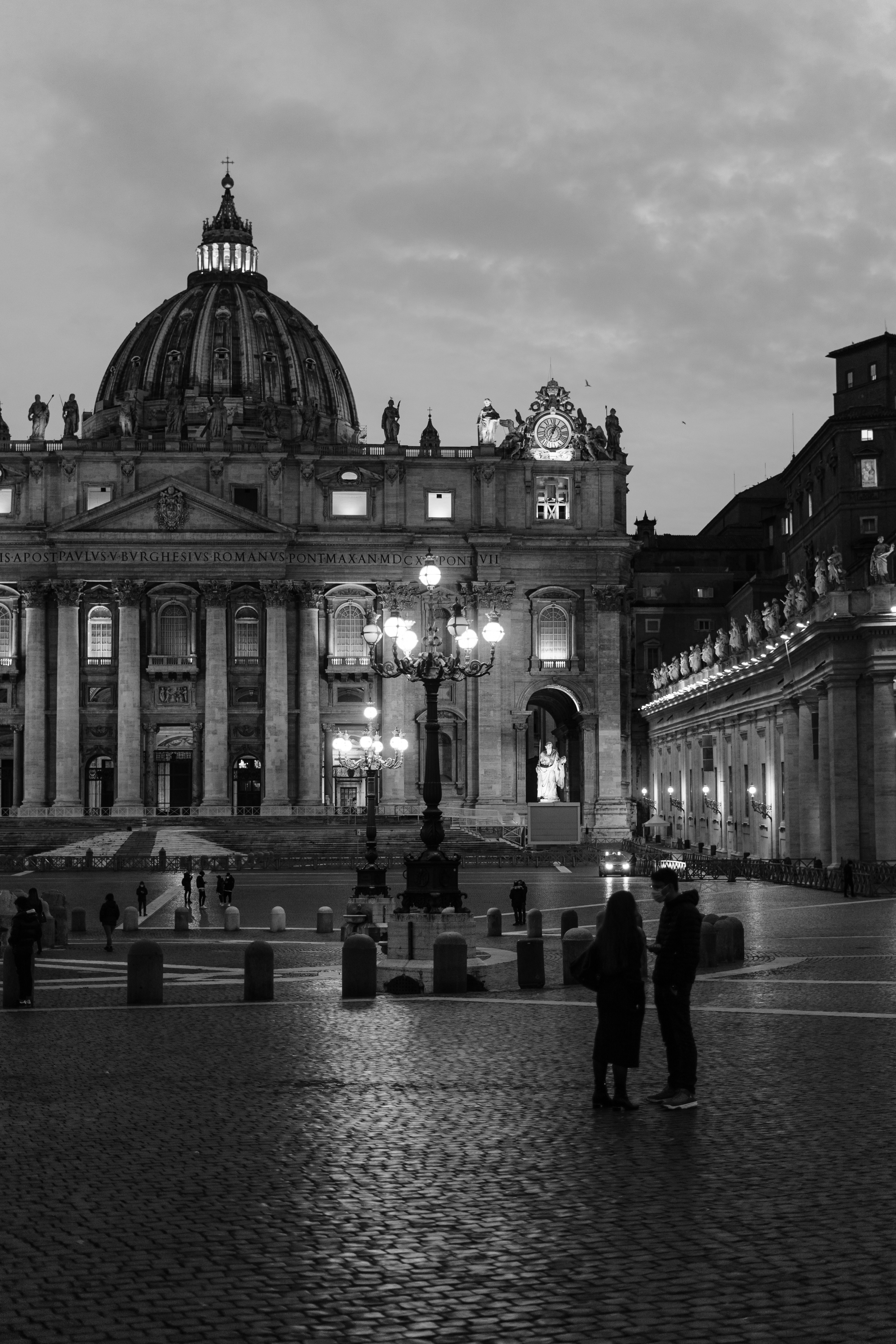 A couple wearing mandatory face masks enjoys St Peter’s Square during blue hour while the city is still in lockdown.  | grayscale photo of people walking near building