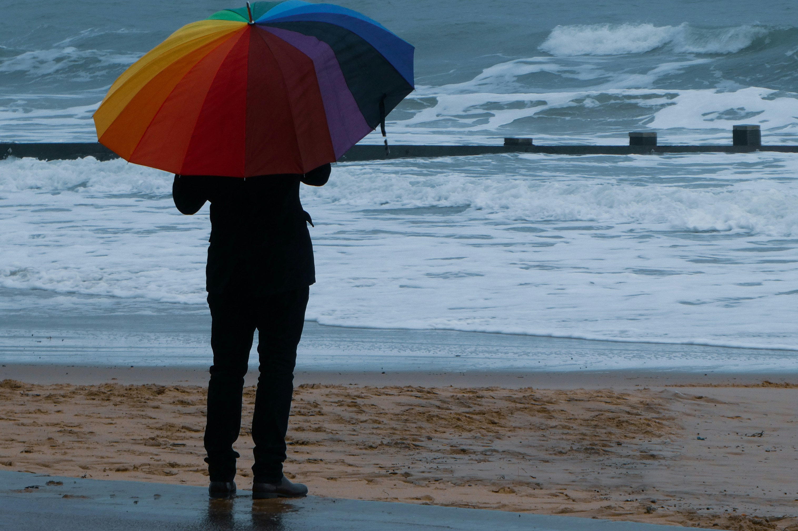 Person holding a vibrant rainbow umbrella while standing on a sandy beach, facing turbulent waves and gray skies.