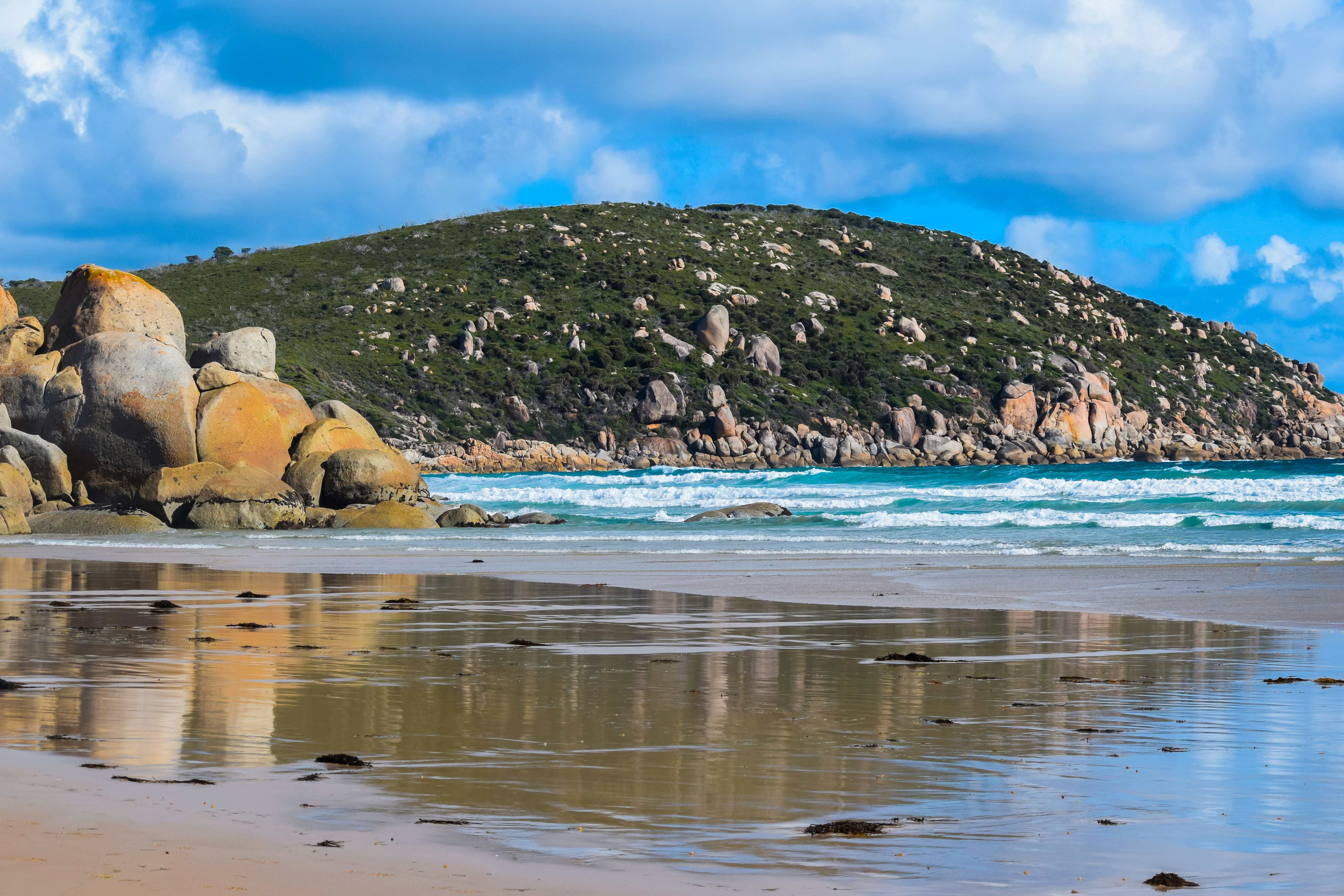 Rocky hill and ocean waves reflected in wet sand under a cloudy sky.
