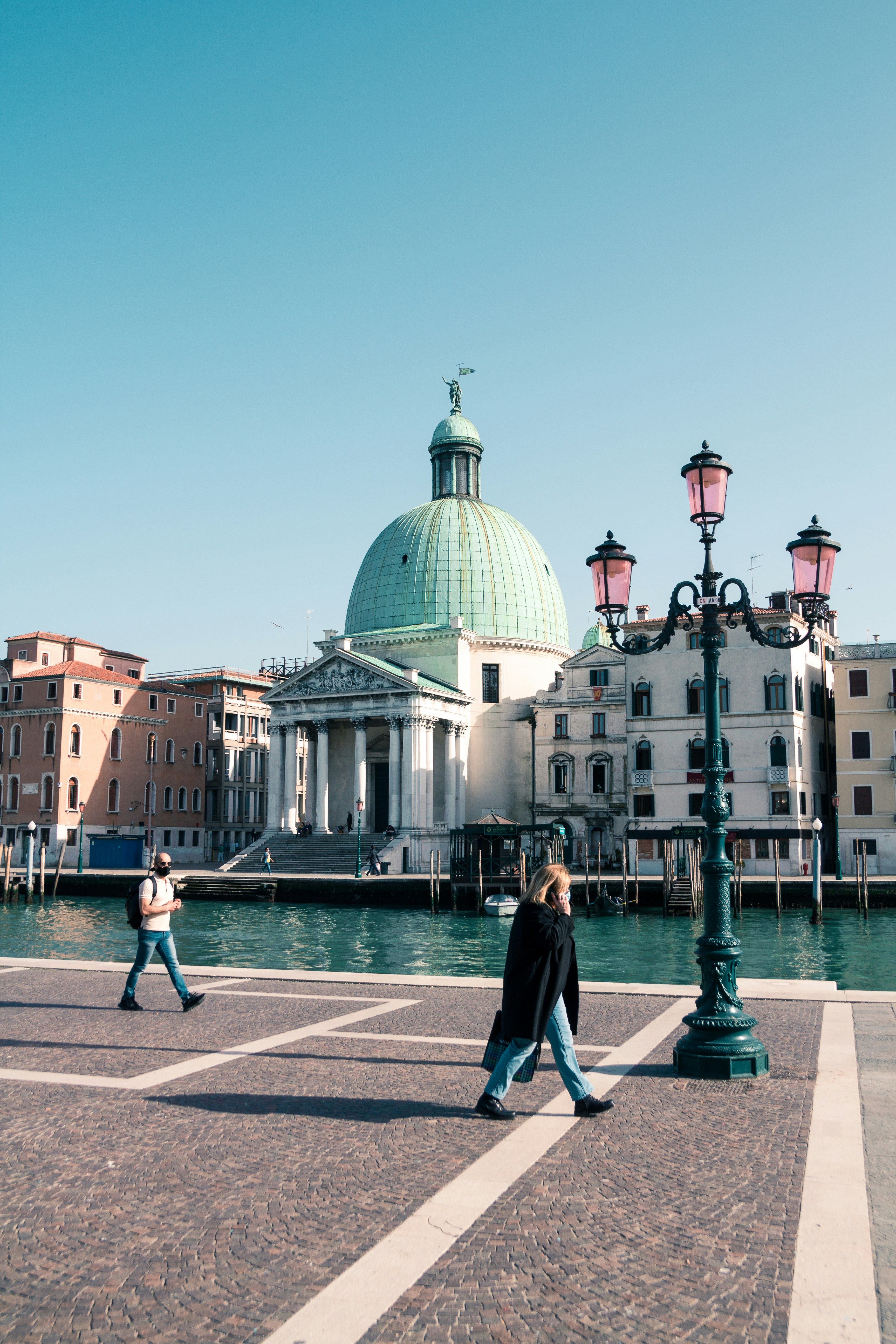 A woman walks along the cobblestone path beside the Grand Canal, with historic buildings and a green-domed structure in the background. The scene captures the essence of Venice's architectural charm.