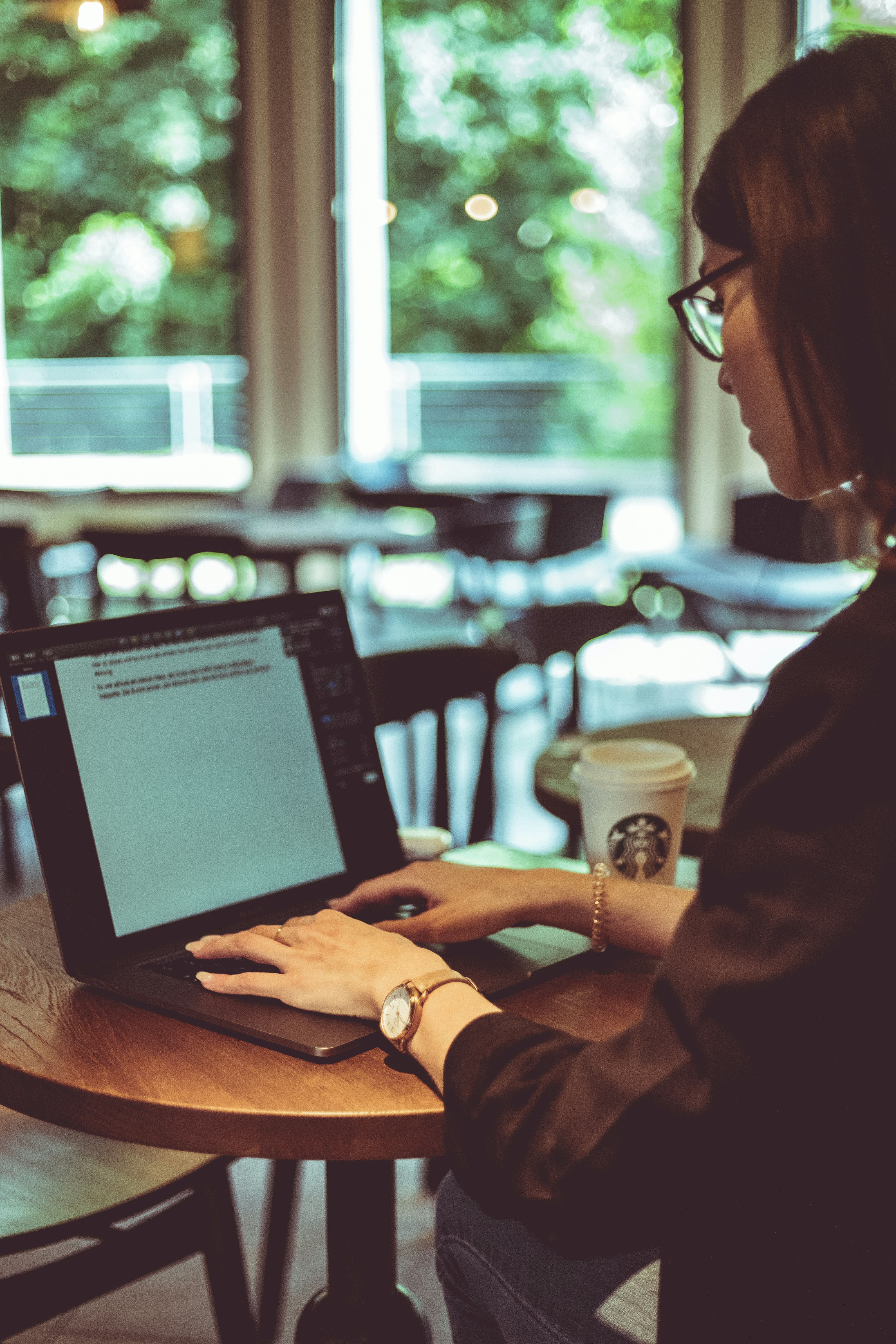 Photograph of a person typing on a laptop at a café table, with a Starbucks cup nearby and warm natural light pouring in through a window.