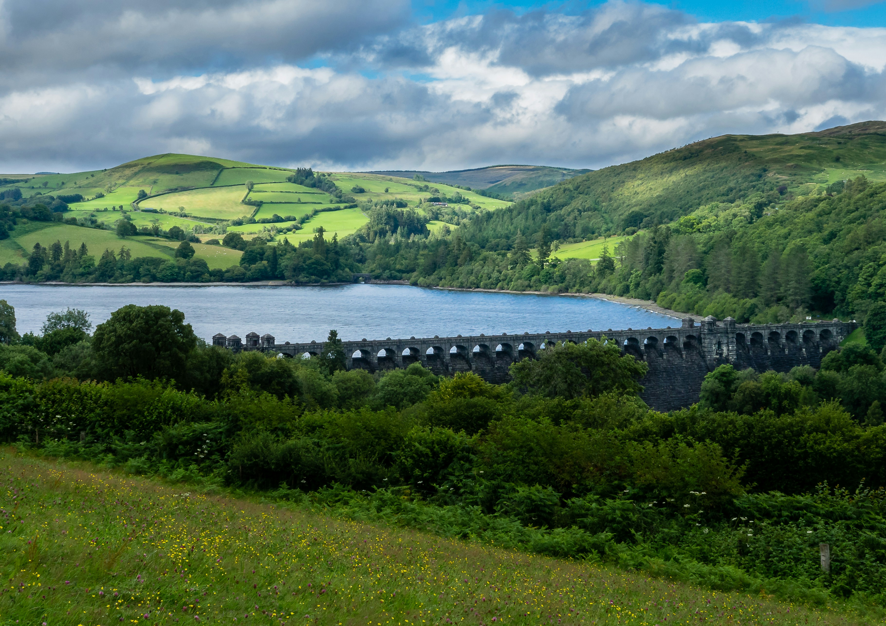 Landscape photograph of a stone viaduct crossing a calm lake, framed by rolling green hills and a cloudy sky.