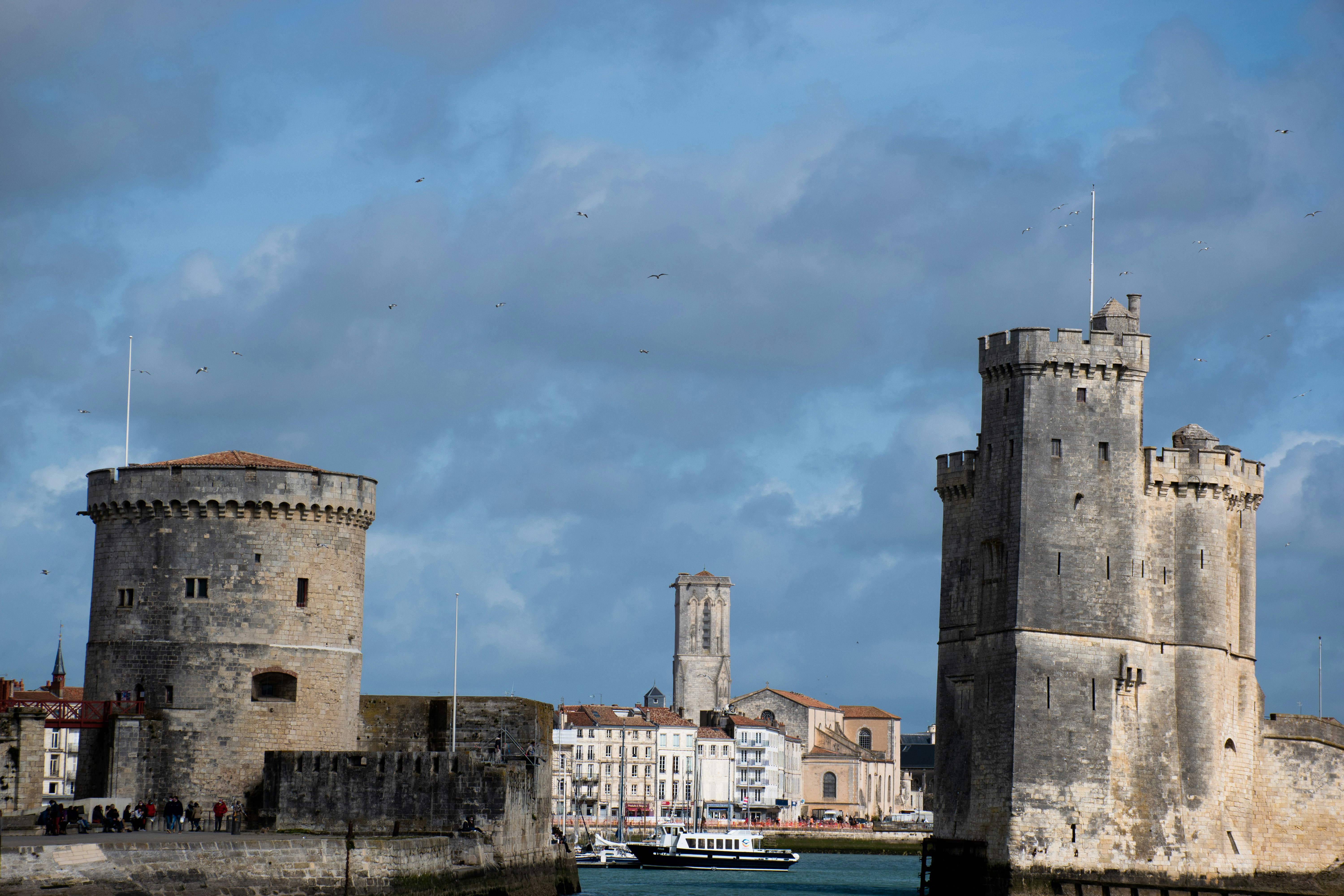 Historic towers stand sentinel by the water's edge, framed by a scenic town backdrop under a dynamic sky.