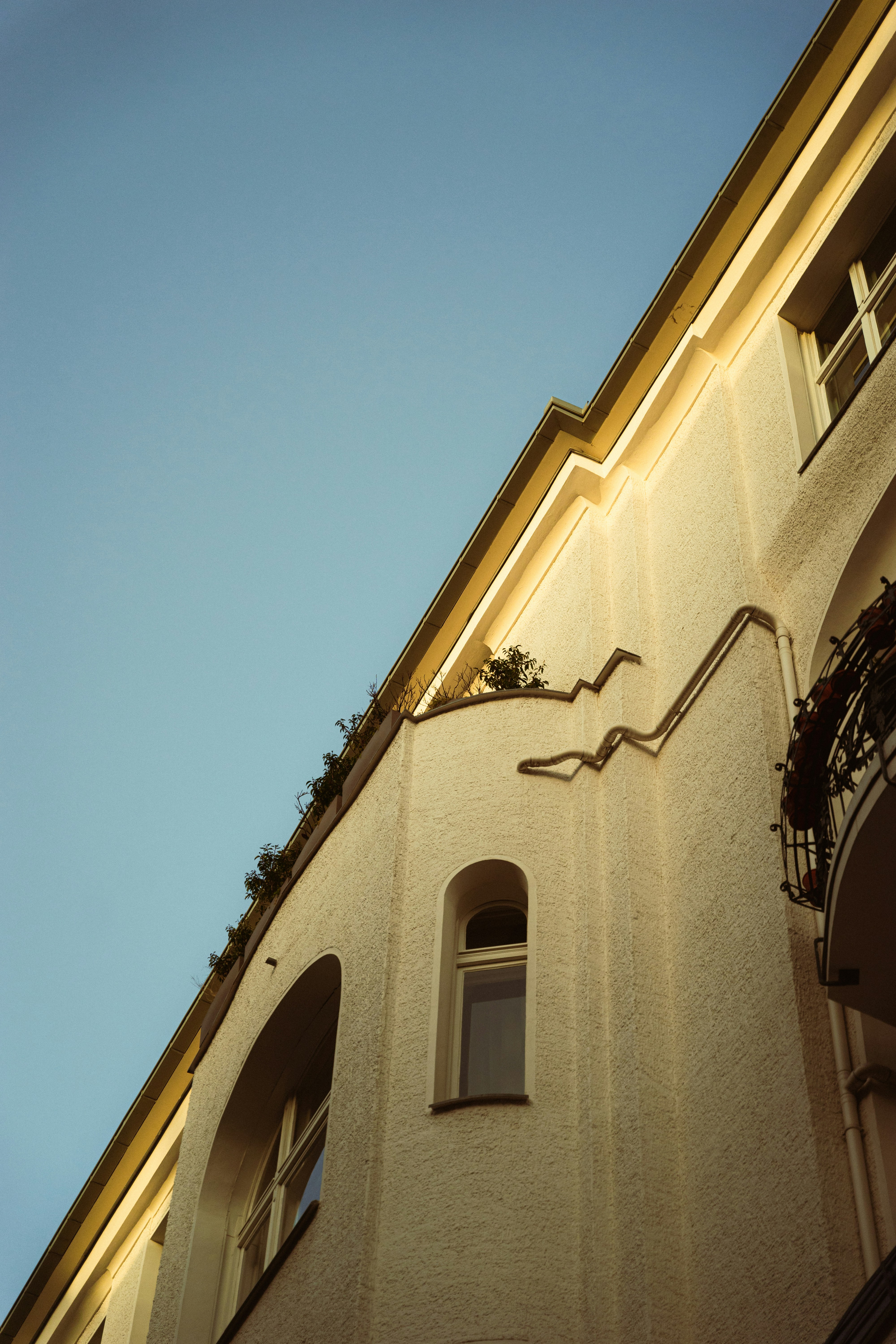 Elegant building facade with soft lighting and greenery, viewed from below against a clear blue sky.
