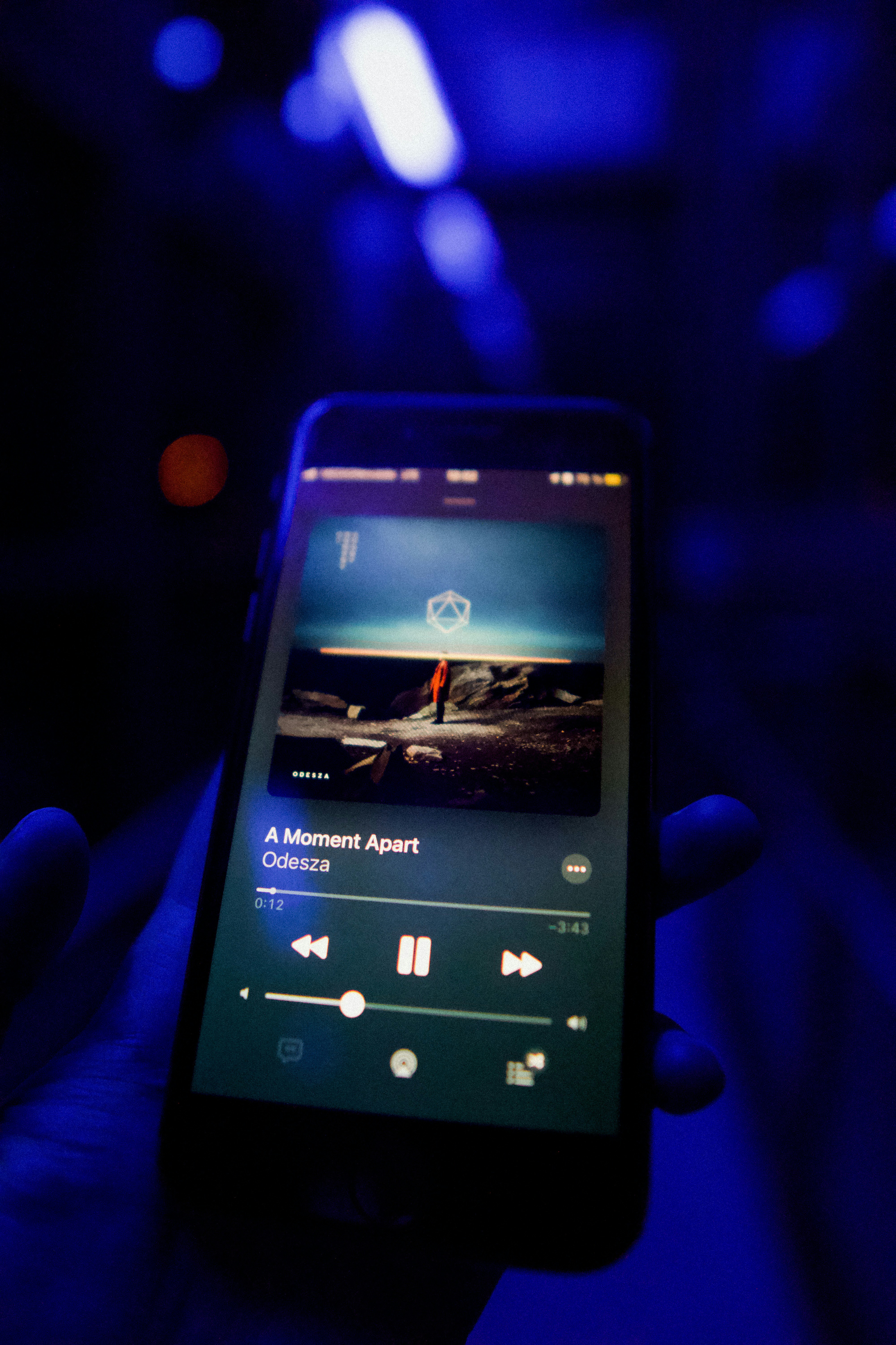A smartphone displaying the album 'A Moment Apart' by Odesza, illuminated by a soft blue light in a dimly lit environment.