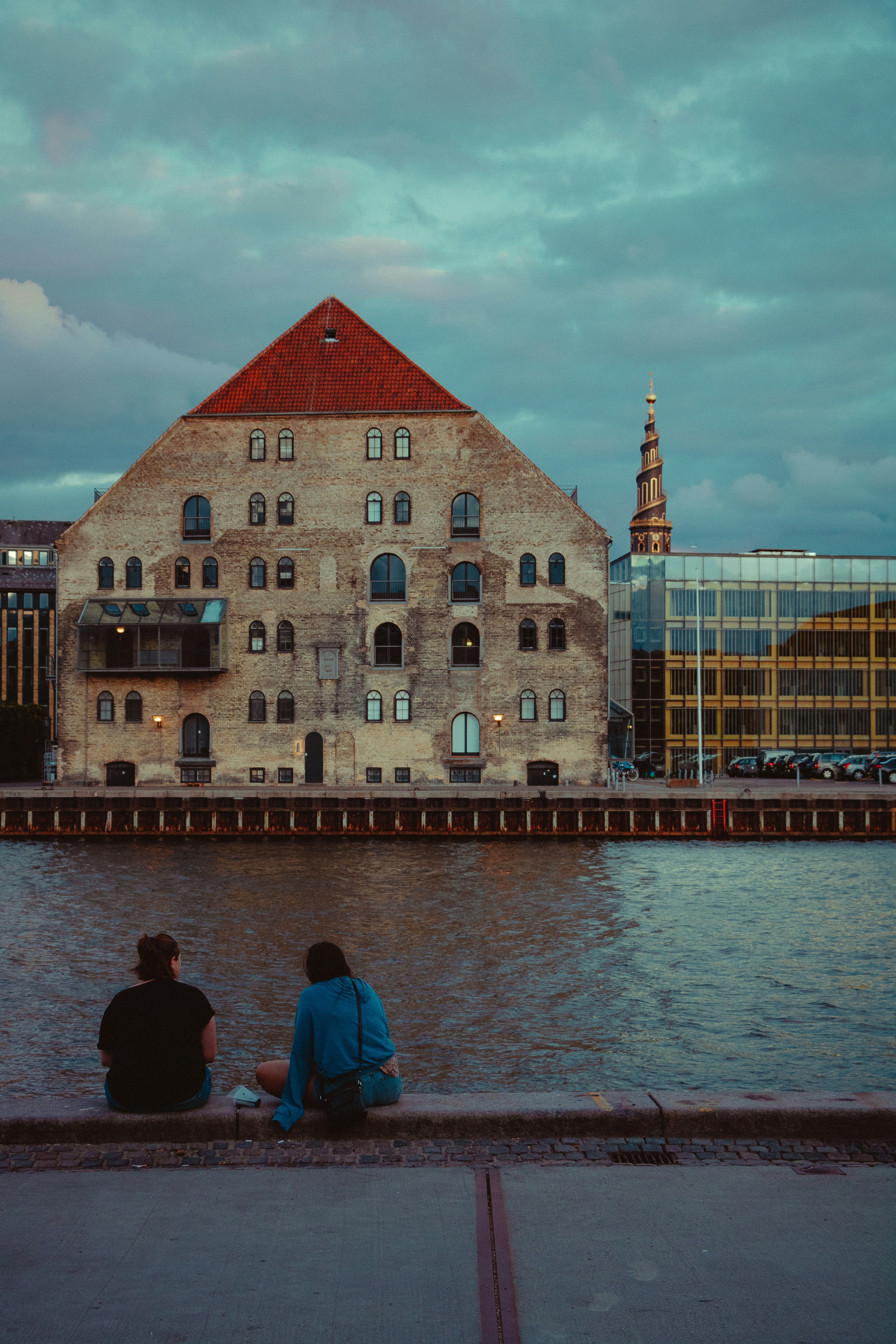 Two individuals seated by the water, gazing at a historic building with unique architecture, under a moody sky.