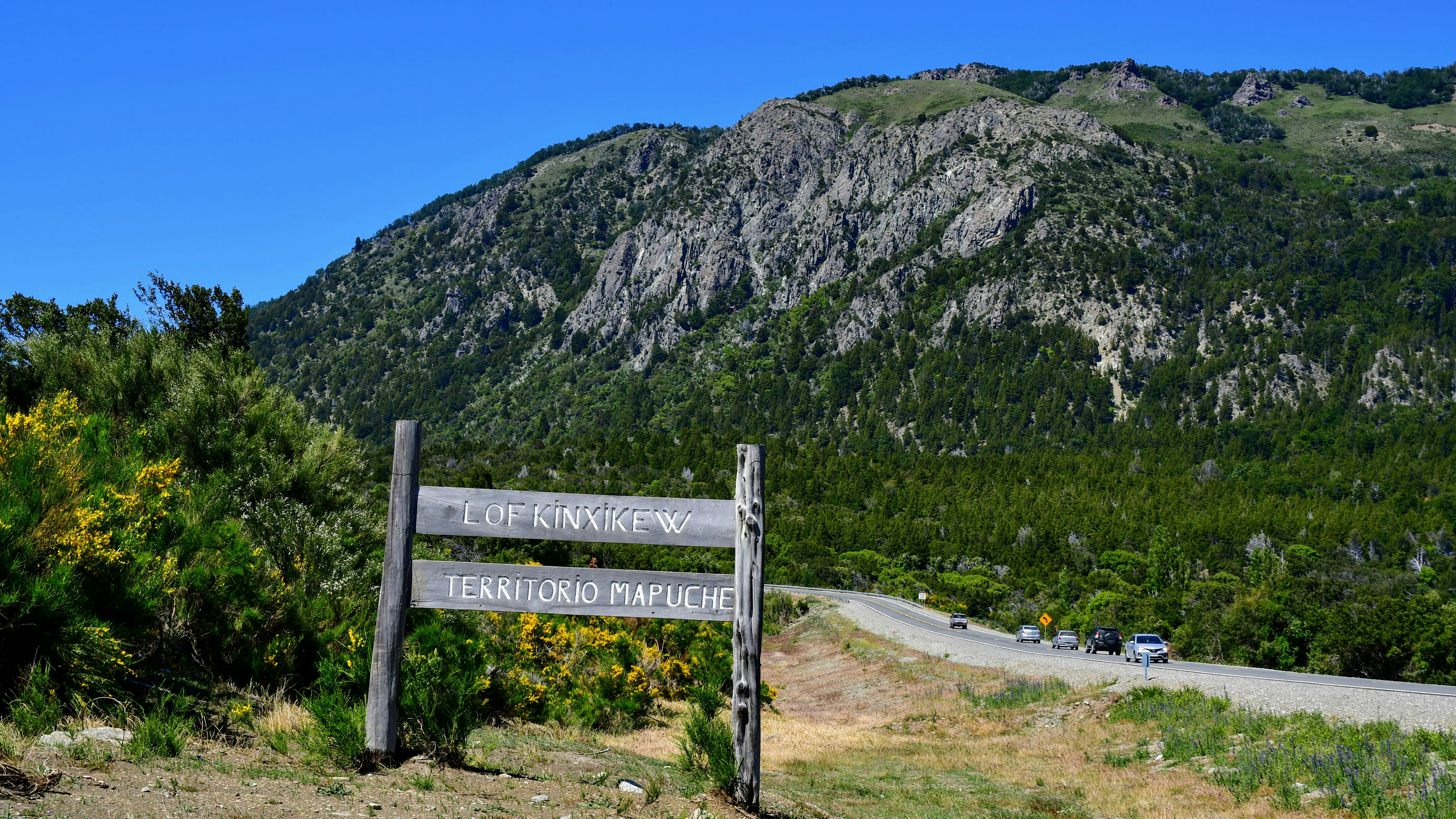 Wooden signpost marks a trailhead leading to a rugged mountain under a clear blue sky.