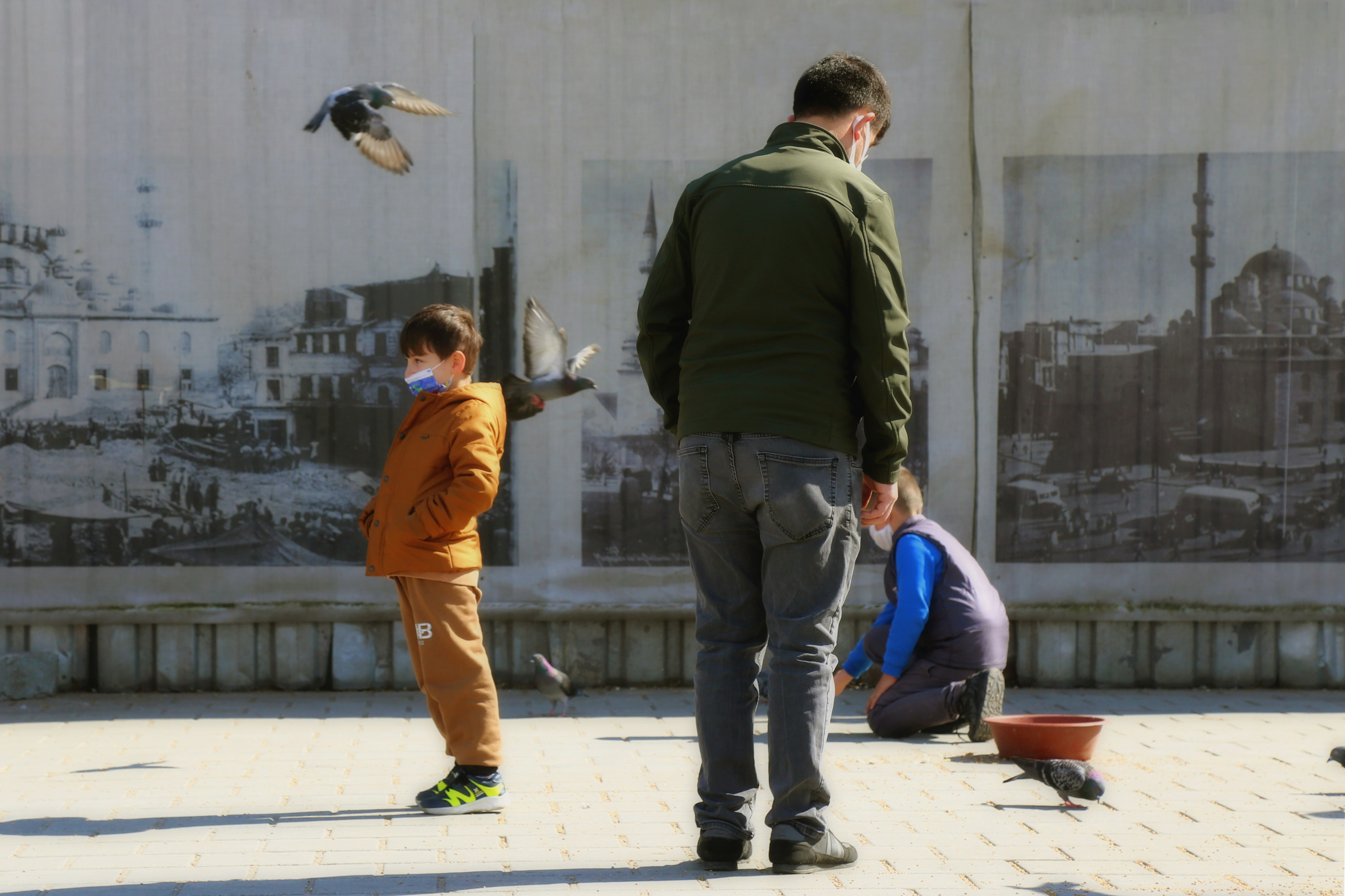 A child in an orange jacket stands still while pigeons flutter around him, capturing a lively scene in a city square.