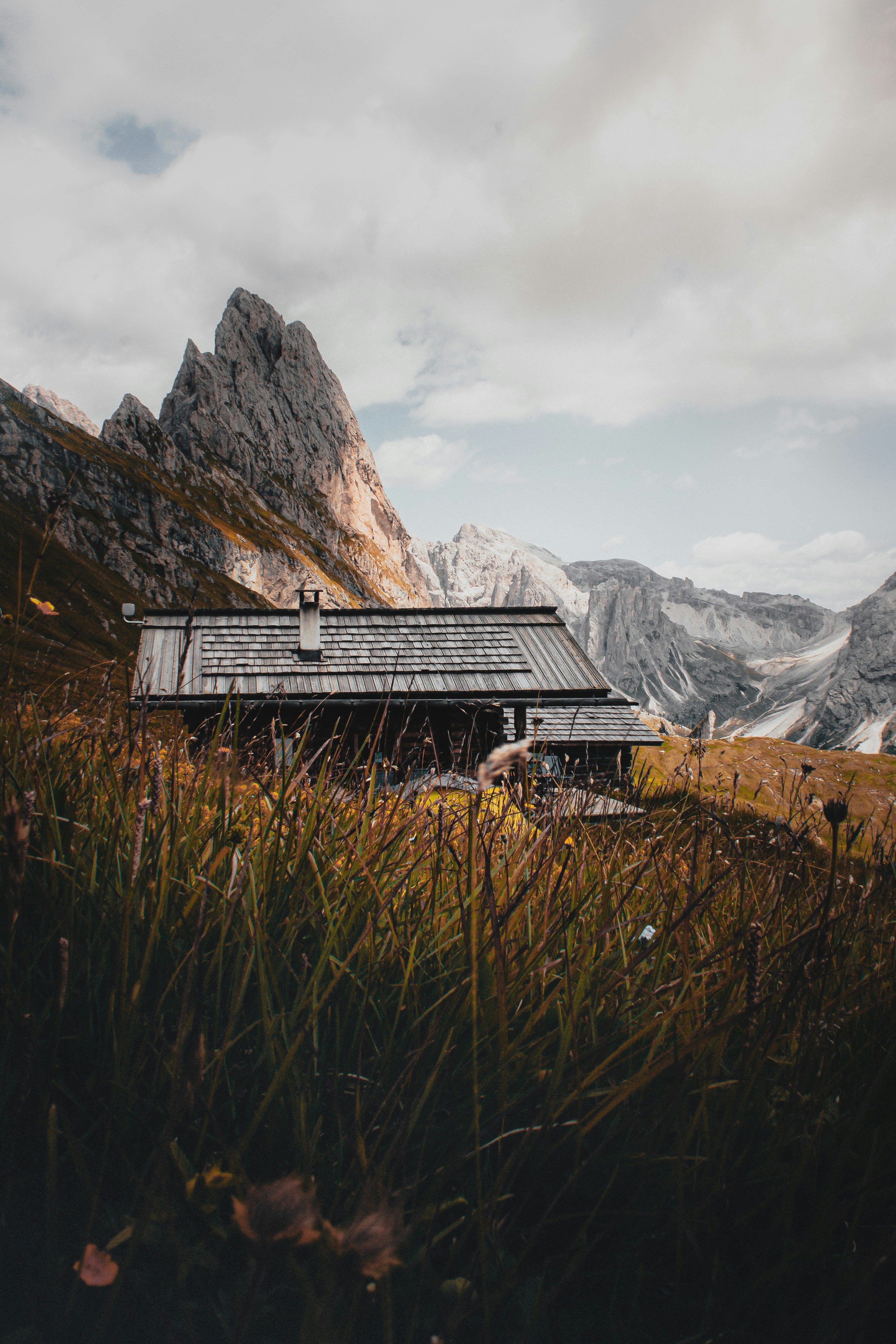 Traditional mountain cabin nestled in lush grass, framed by towering peaks under a cloudy sky.