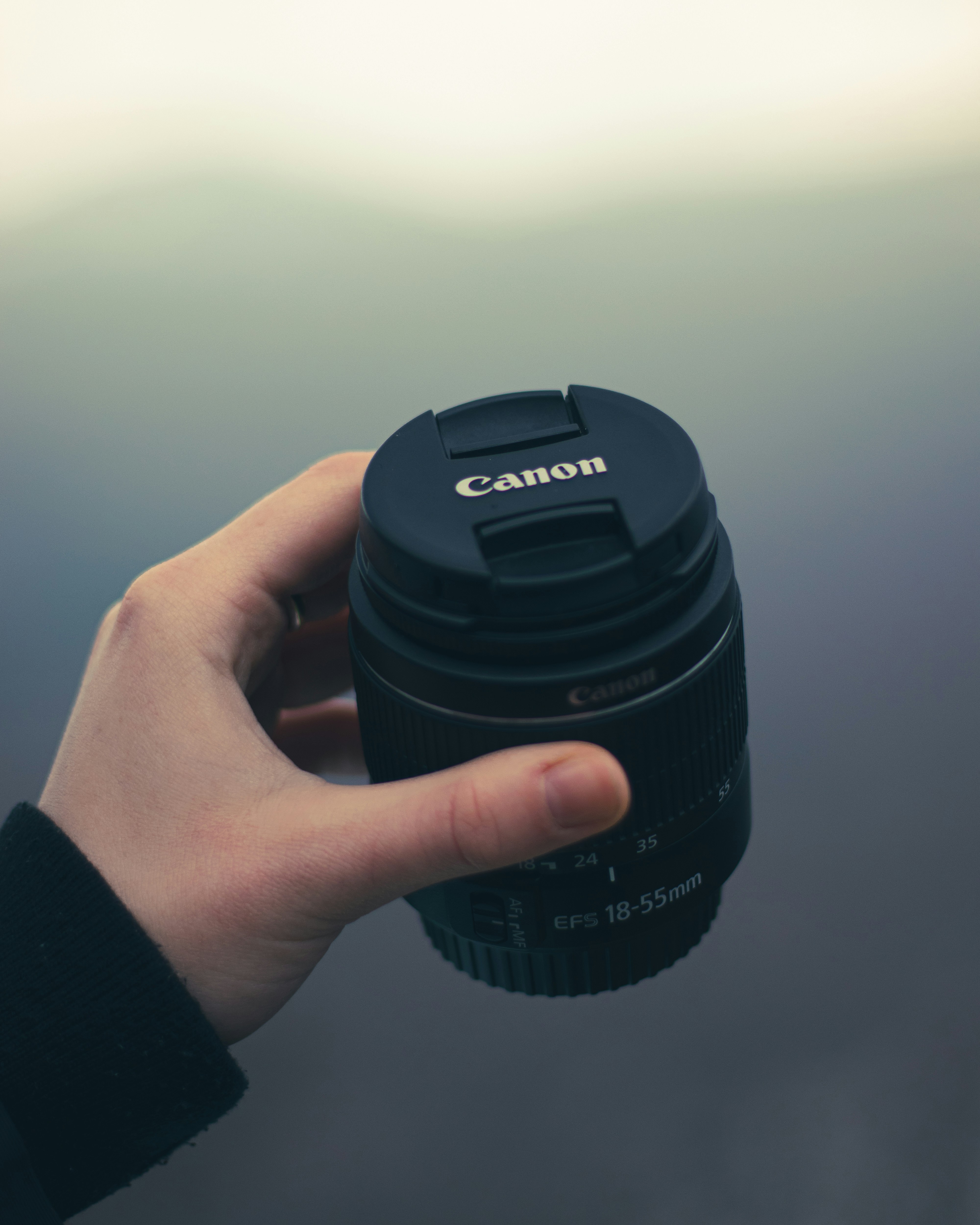 Hand holding a Canon EF-S 18-55mm lens against a blurred background, emphasizing the connection between photographer and equipment.