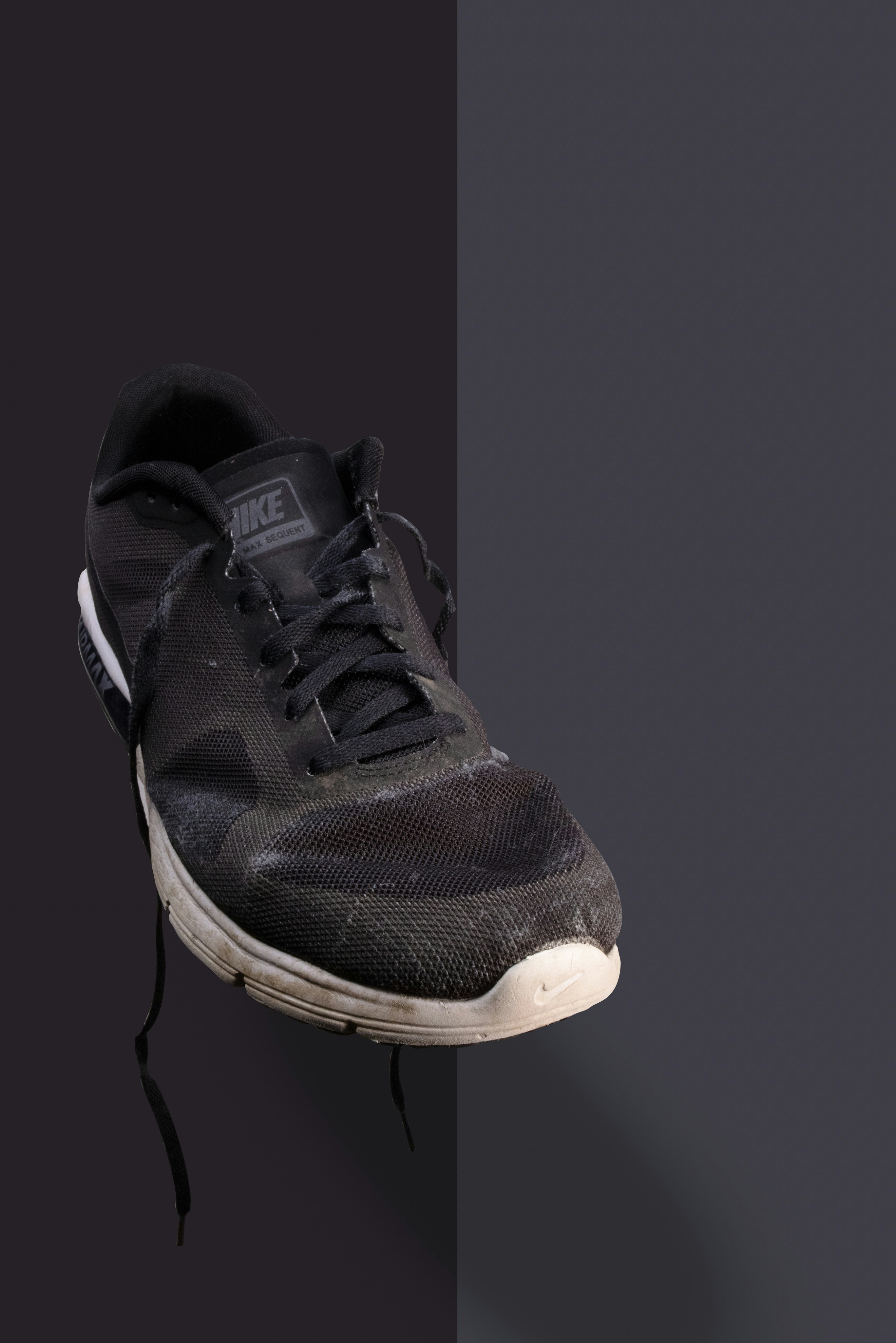 A black Nike sneaker suspended against a contrasting background, showcasing its worn-out condition and intricate details. The focus is on the shoe's texture and form.