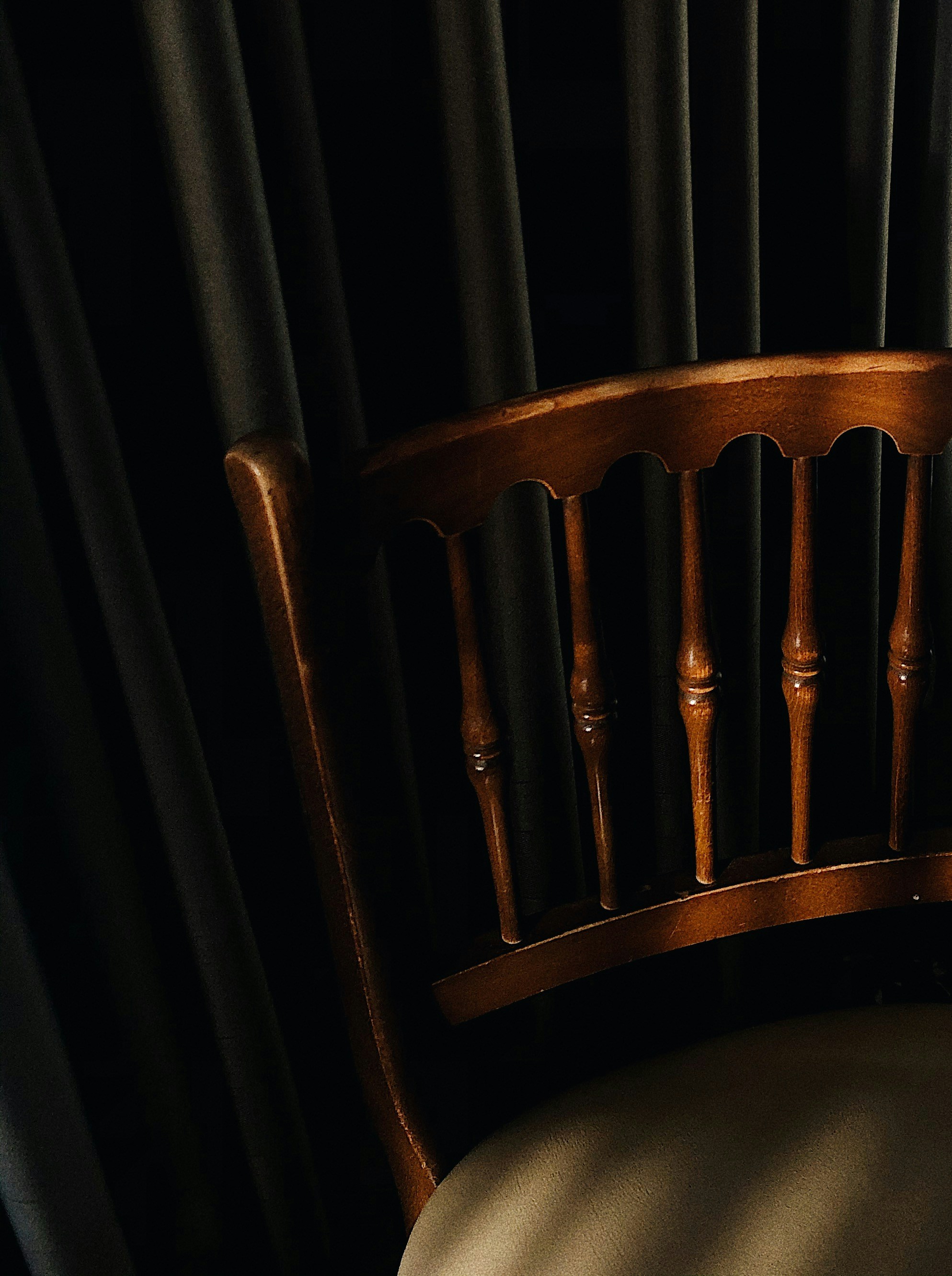 A wooden chair partially illuminated against dark curtains, highlighting its intricate design and texture.