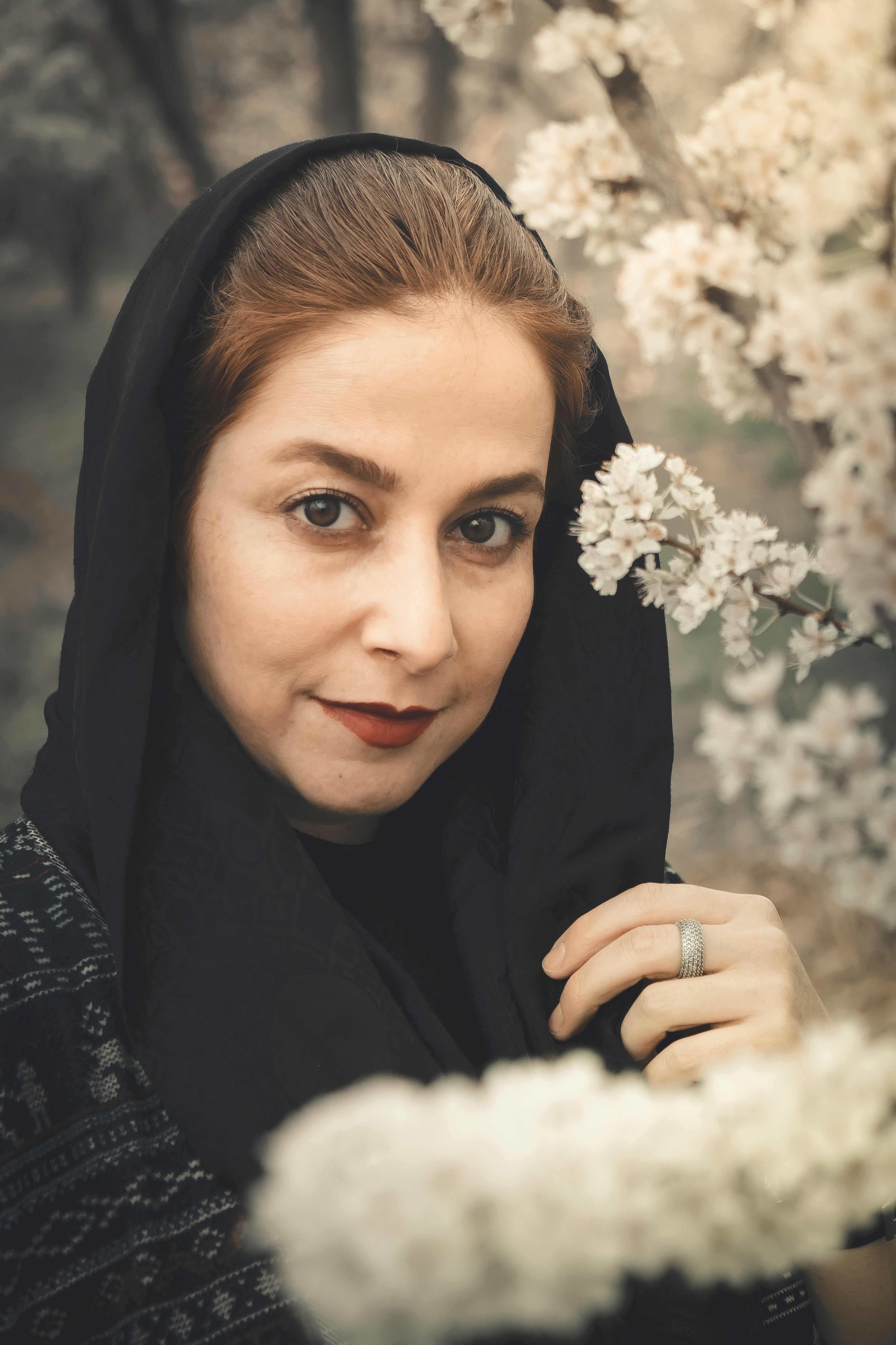 A woman in a black hijab gazes softly at the camera, framed by delicate white blossoms in a serene setting.