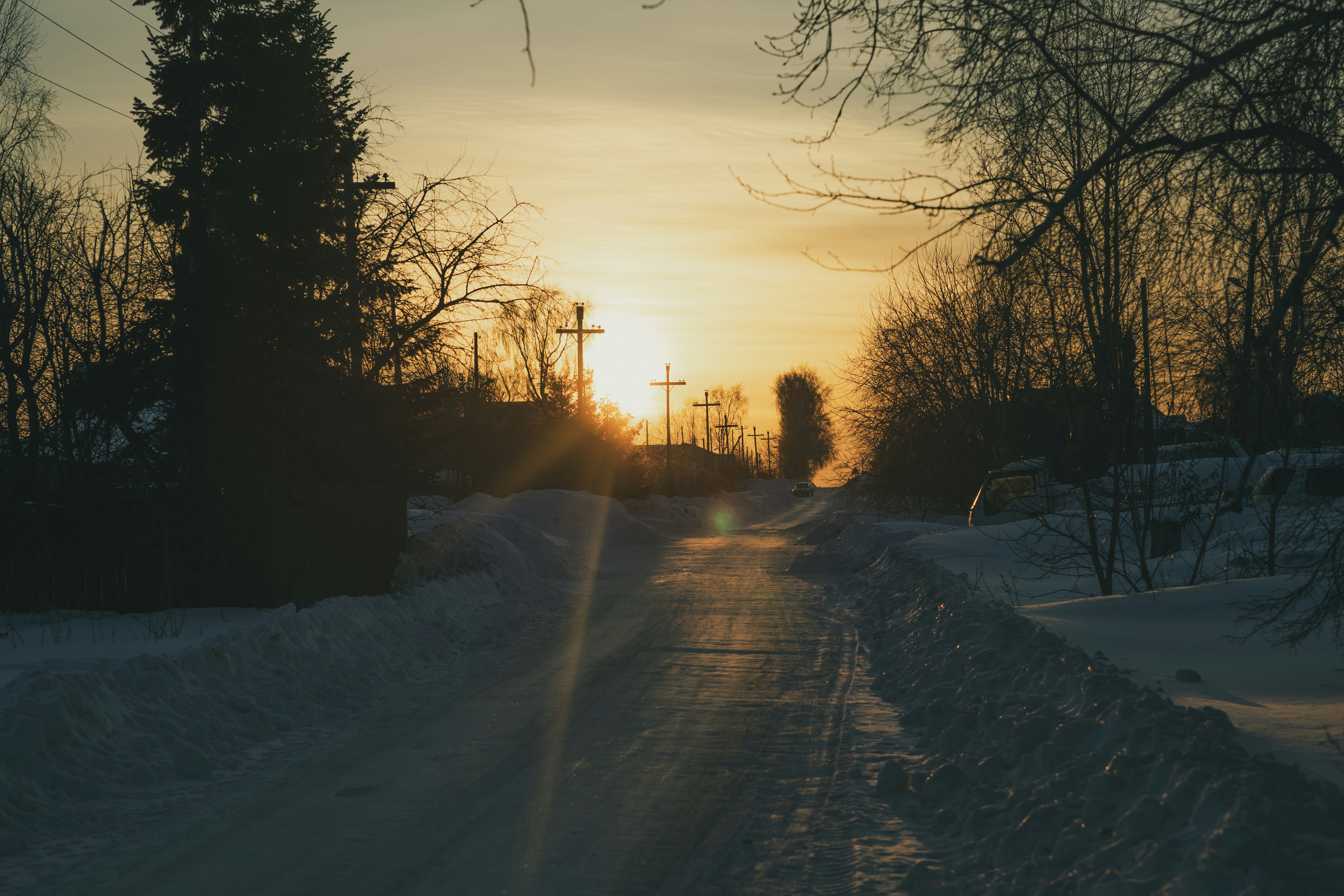 Golden sunset illuminating a snow-covered road flanked by bare trees, with a silhouette of a cross in the distance.