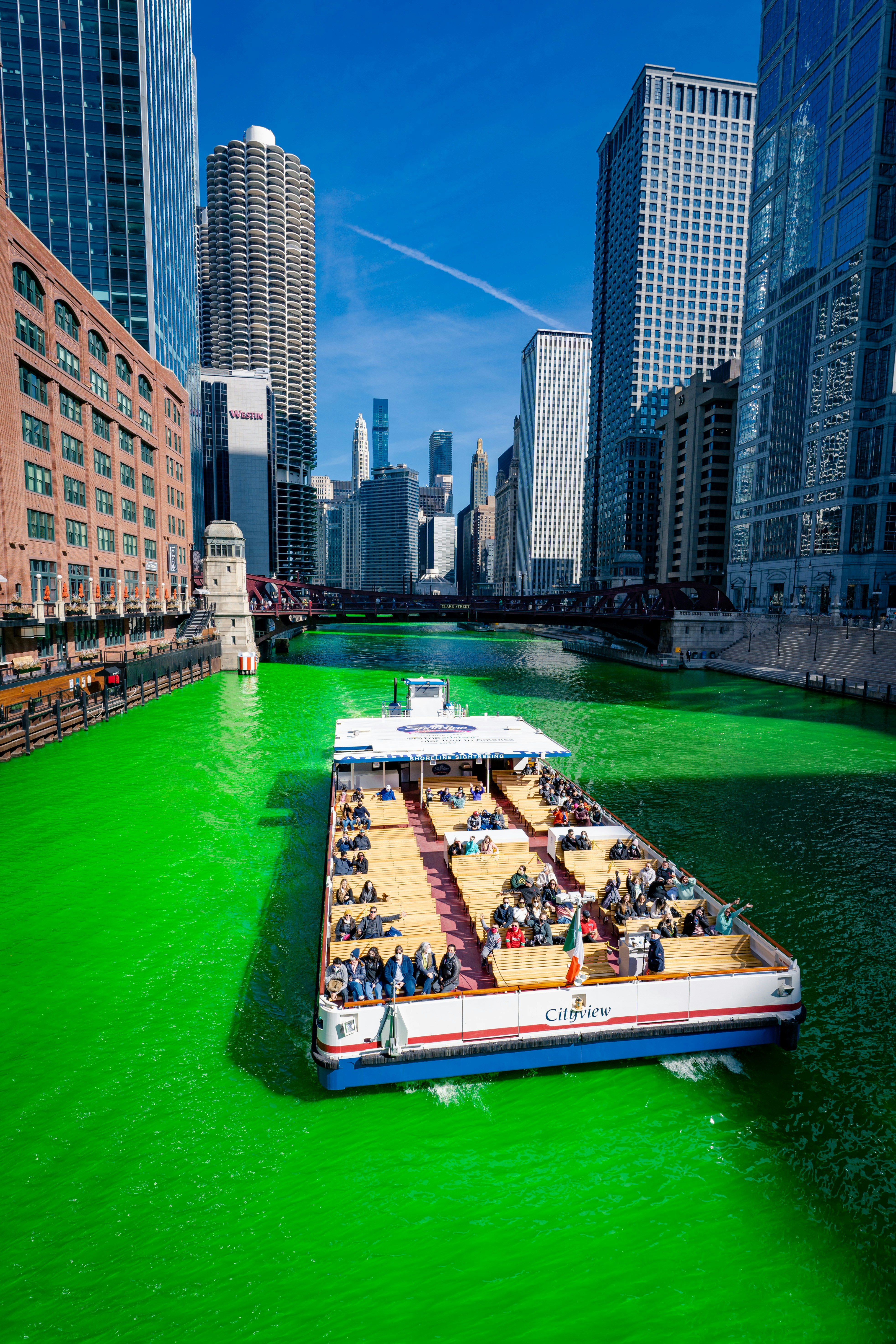 A vibrant green river flows through a bustling cityscape, with a sightseeing boat filled with passengers enjoying the view. Skyscrapers rise majestically on either side.