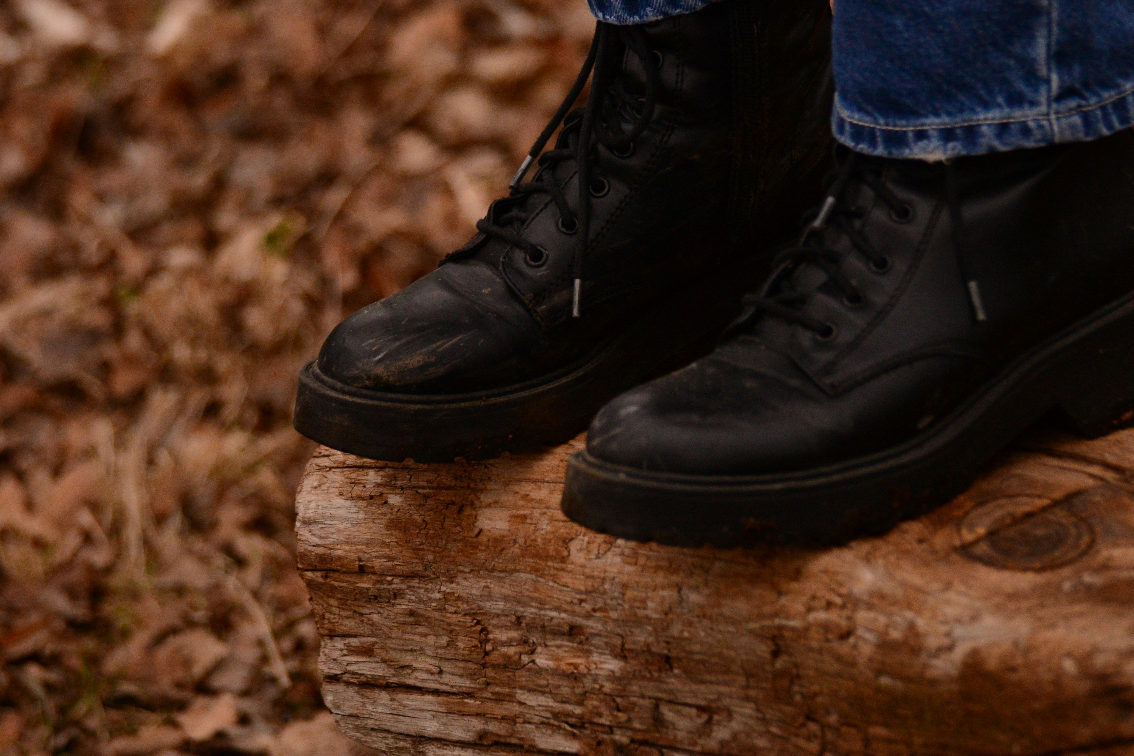 Close-up of rugged black boots resting on a weathered log amidst a backdrop of fallen leaves.
