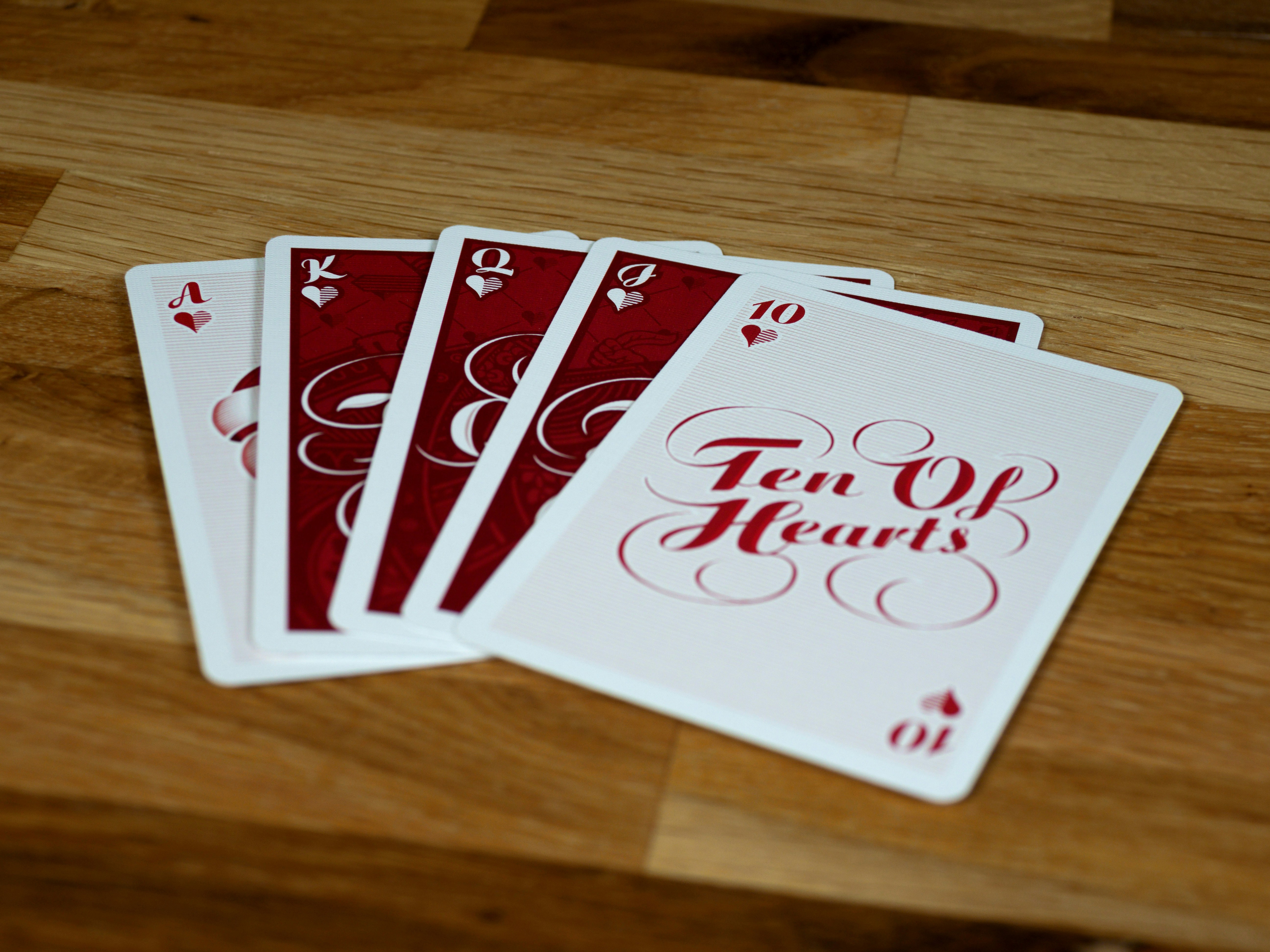 A hand of playing cards featuring the Ten of Hearts, flanked by the King, Queen, and Ace of Hearts. The cards are spread out on a wooden surface.