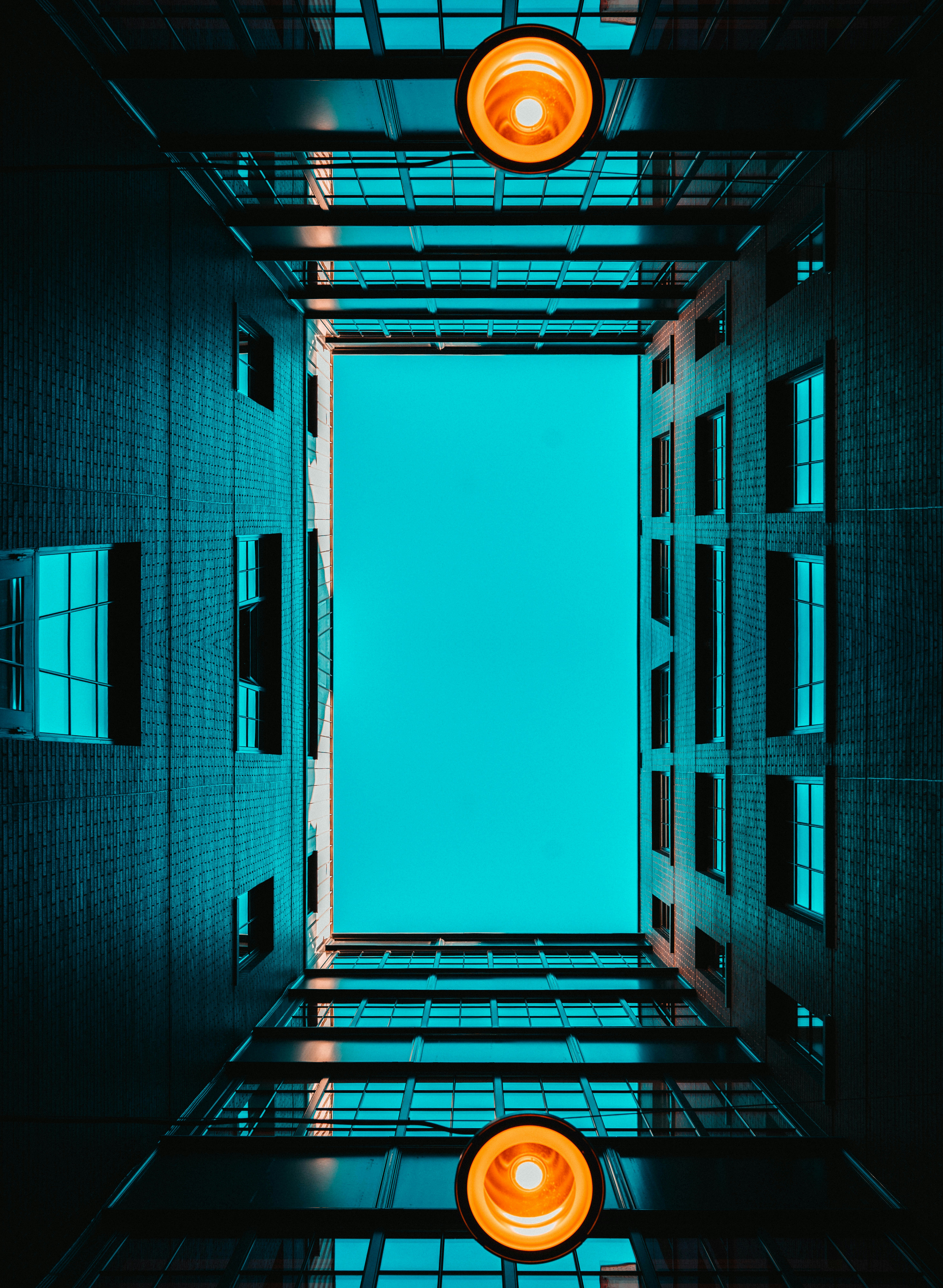 Looking up through a modern architectural courtyard, vibrant blue sky contrasts with the surrounding brick and glass structures, illuminated by circular lights.