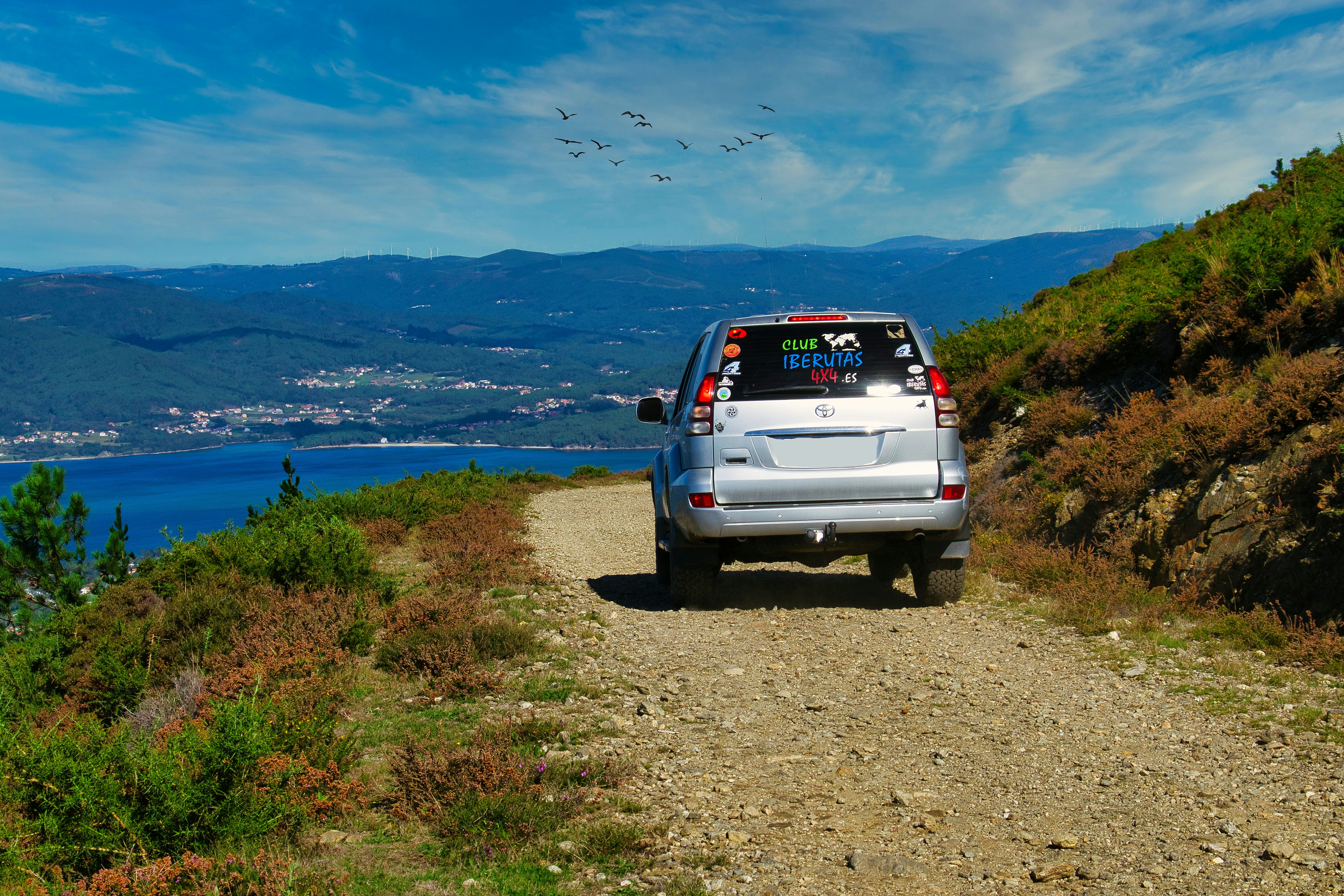 SUV navigating a rugged path overlooking a serene coastal landscape dotted with greenery and distant hills.