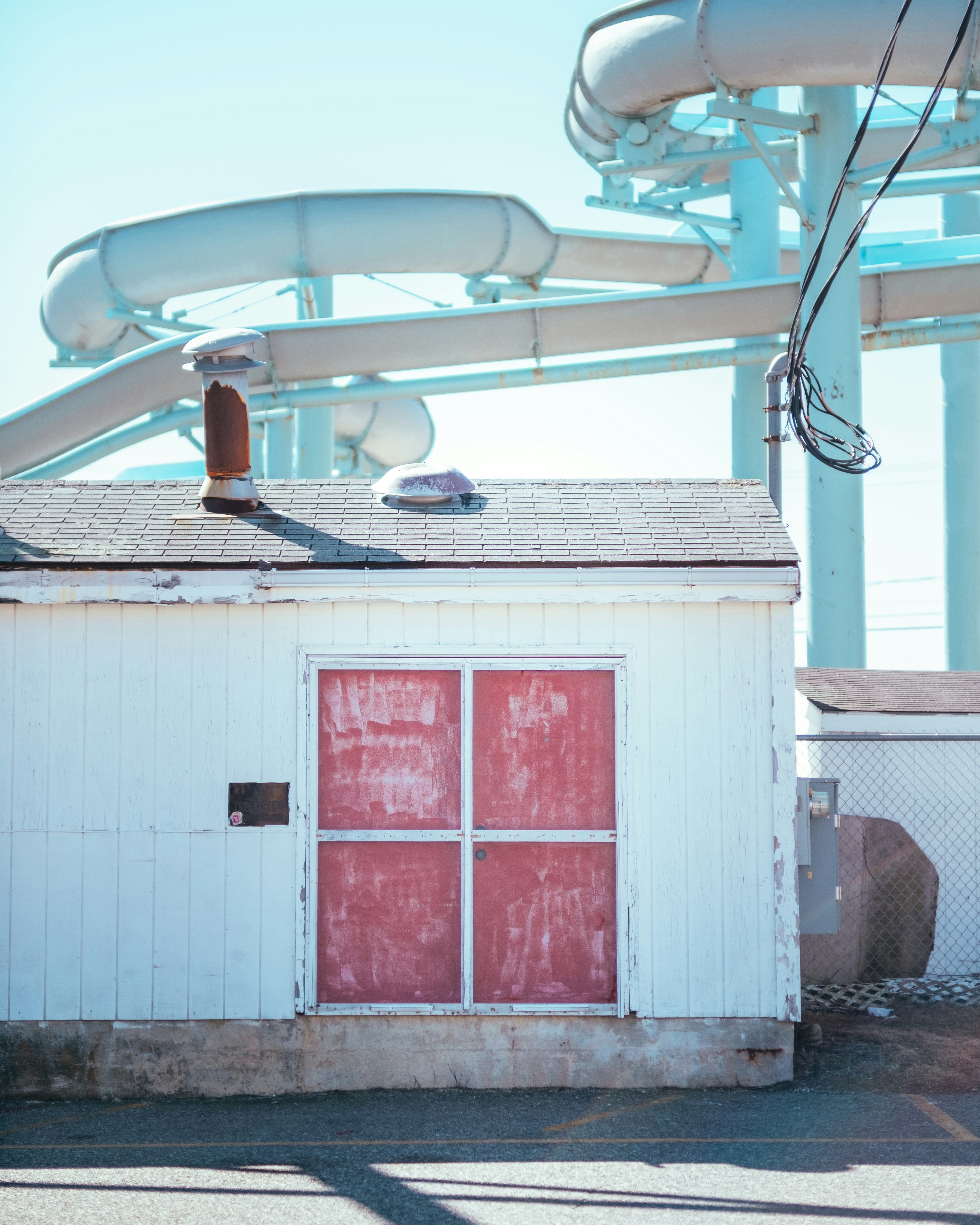 A weathered white shed with a red door stands in the foreground, juxtaposed against a towering aqua waterslide structure in the background.