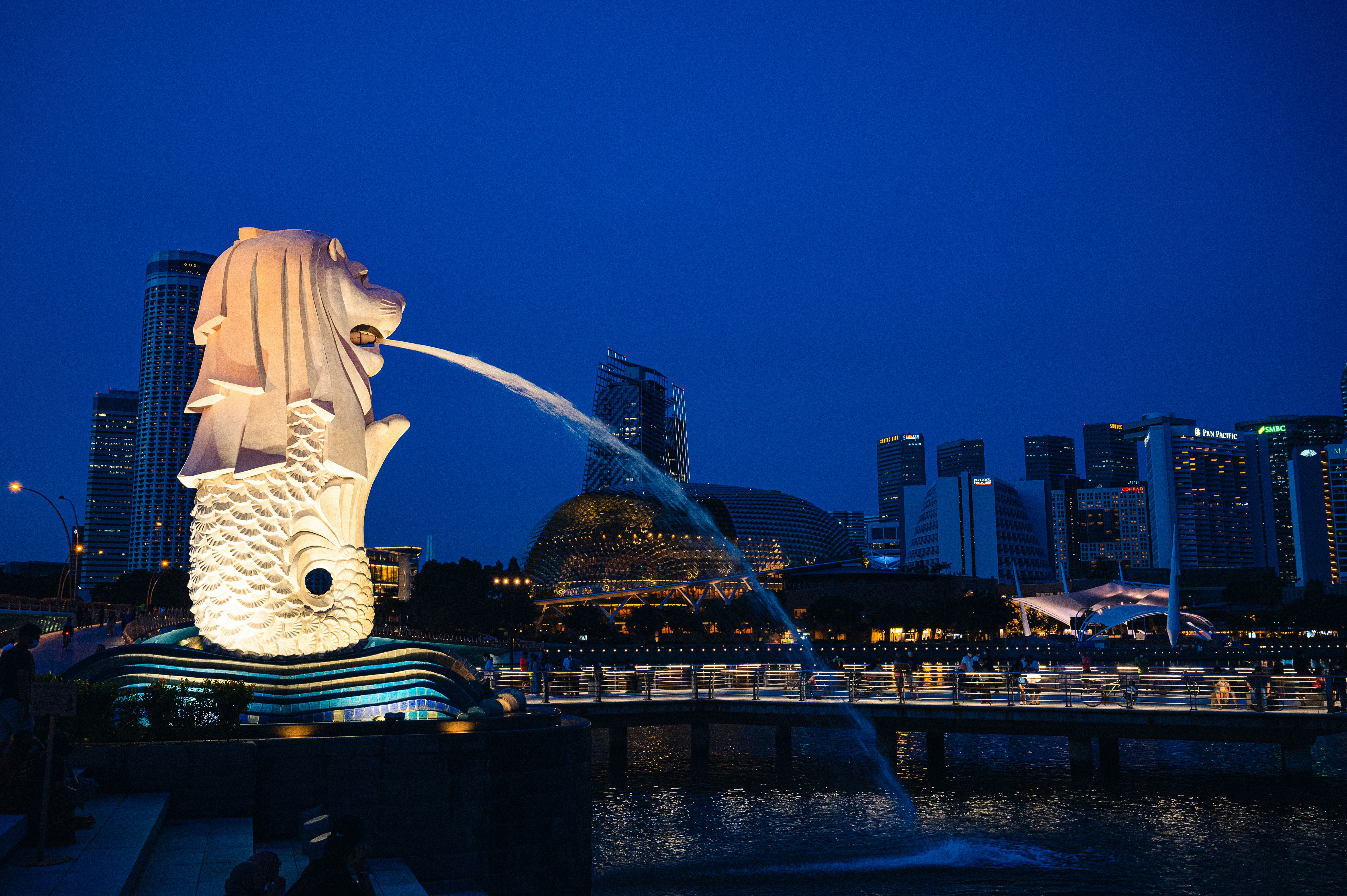 Merlion statue with water spout at dusk, set against a cityscape with illuminated buildings.