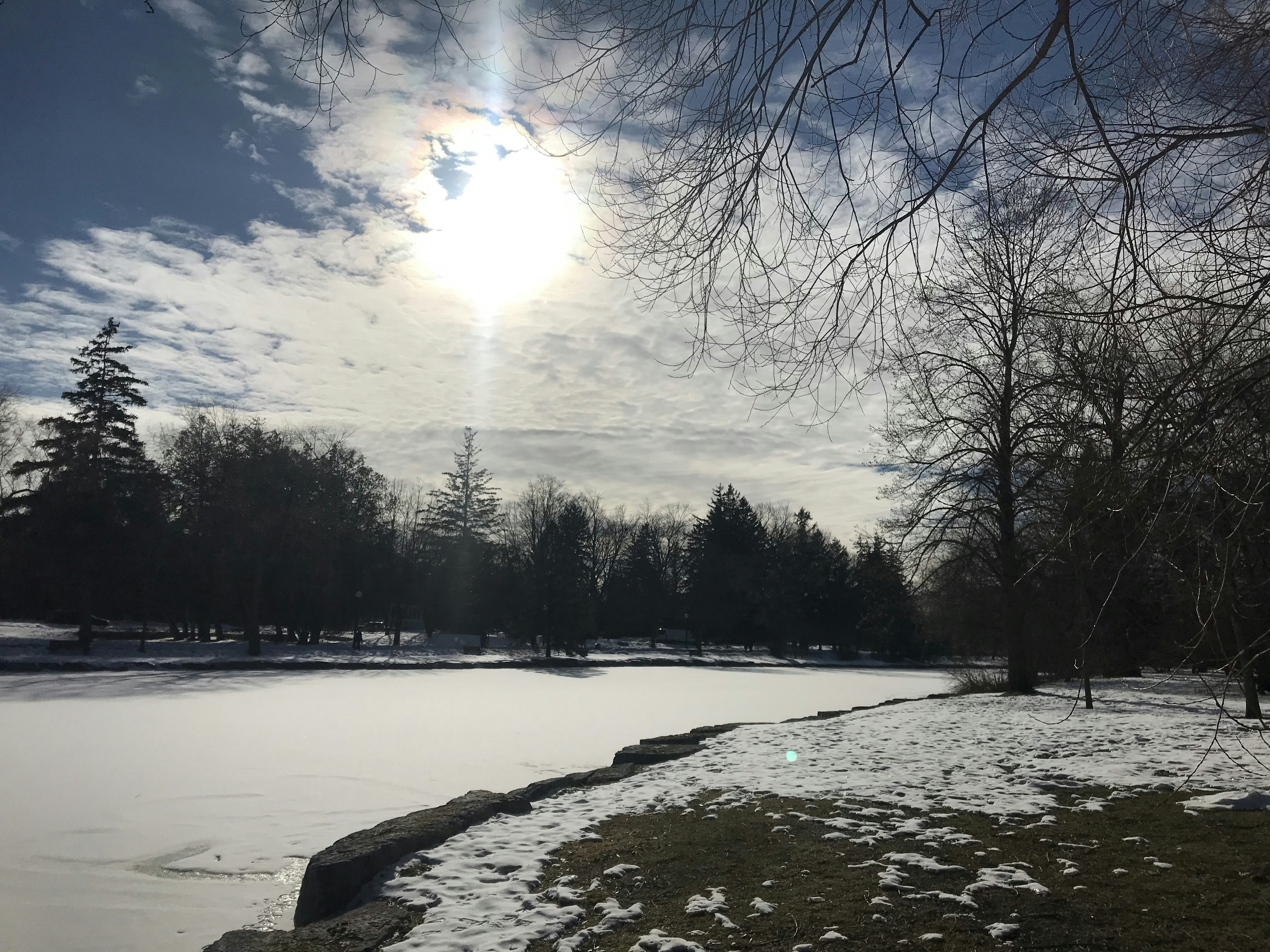 Bright sun peeking through clouds over a snow-covered landscape with a frozen pond and bare trees. The scene captures the tranquility of winter.