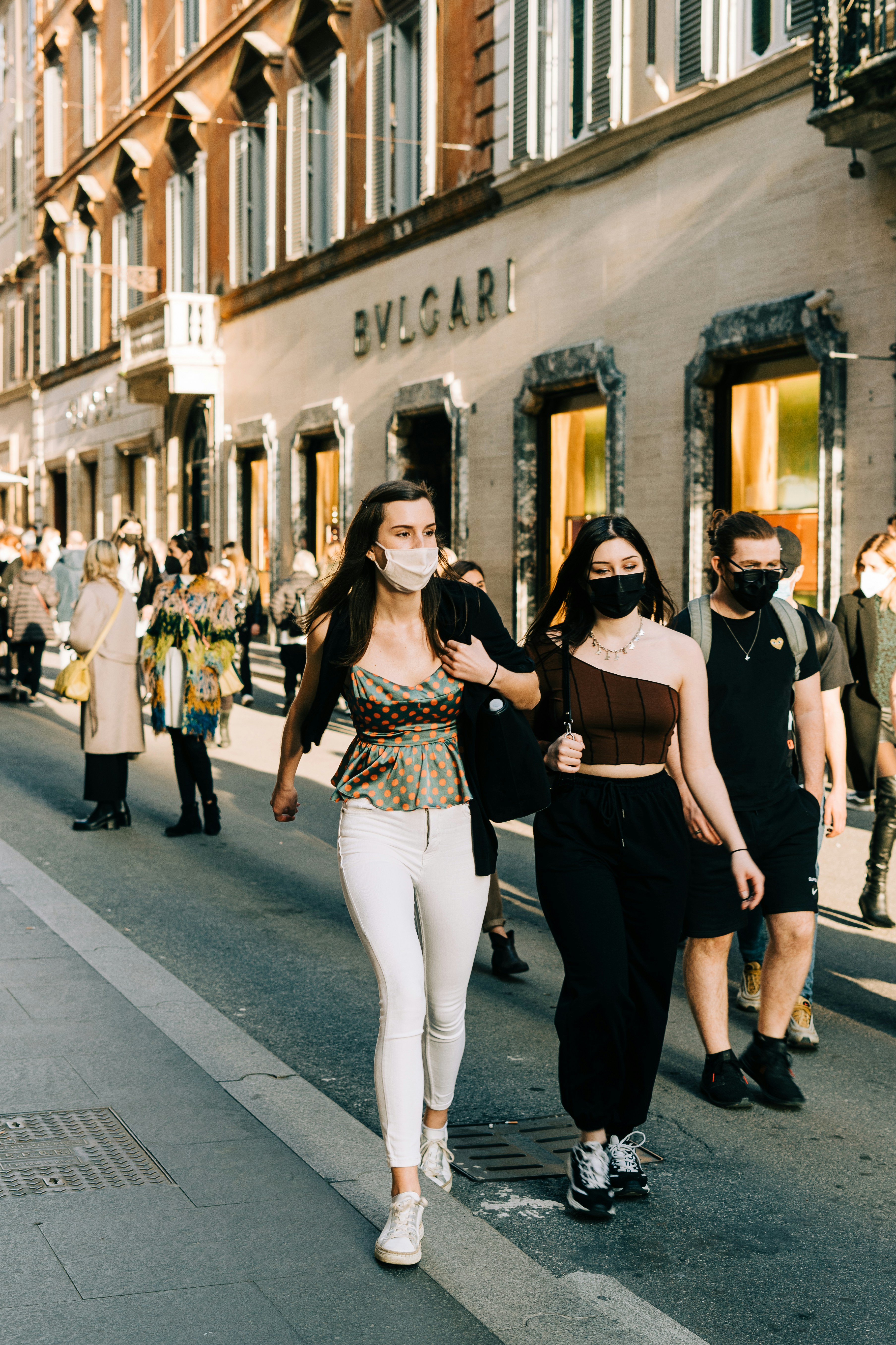 Two women walk confidently down a bustling street, showcasing contemporary fashion against a backdrop of luxury storefronts. The scene captures the essence of urban life post-pandemic.