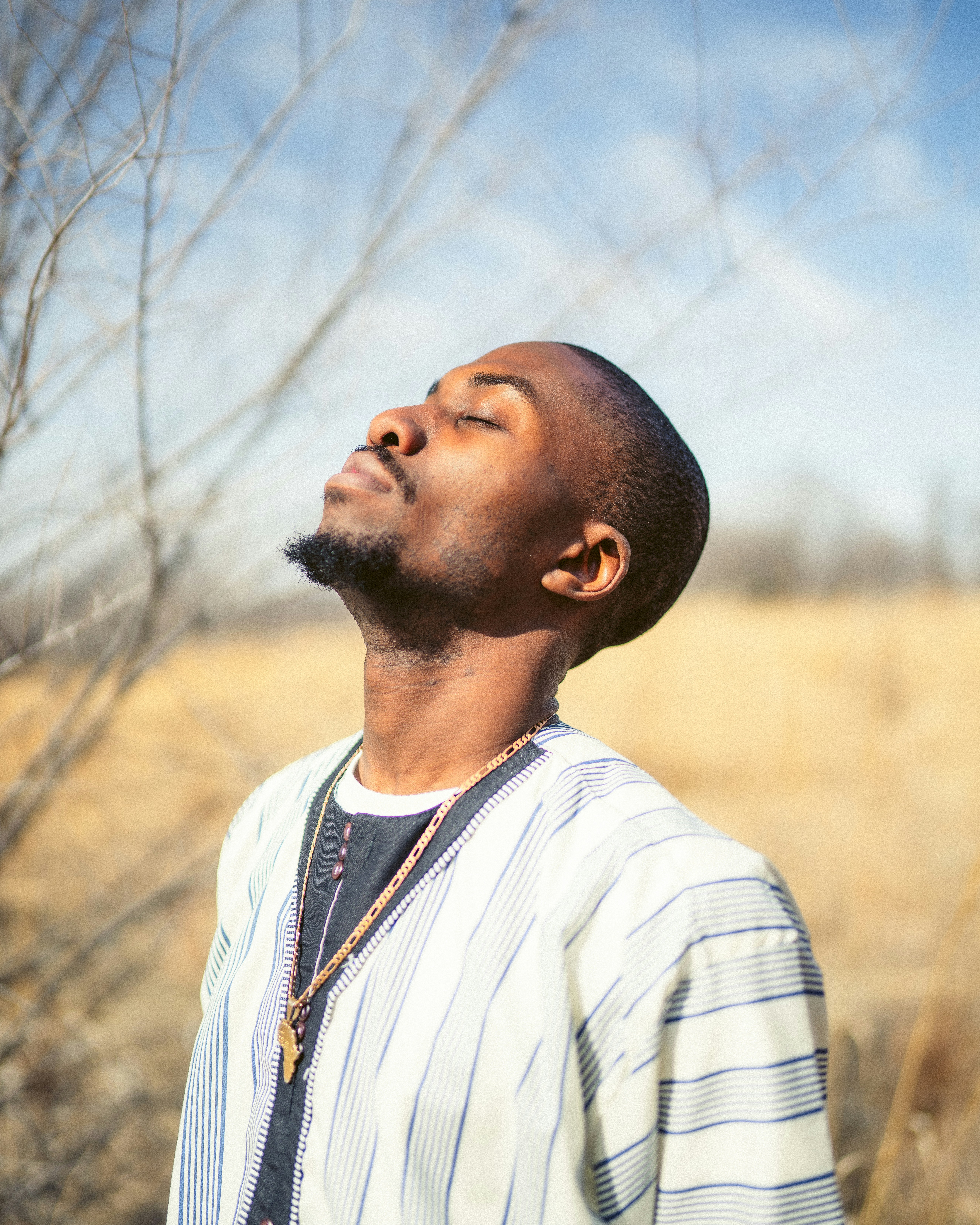 Man with closed eyes and serene expression stands amidst dry grass and sparse branches, basking in sunlight.