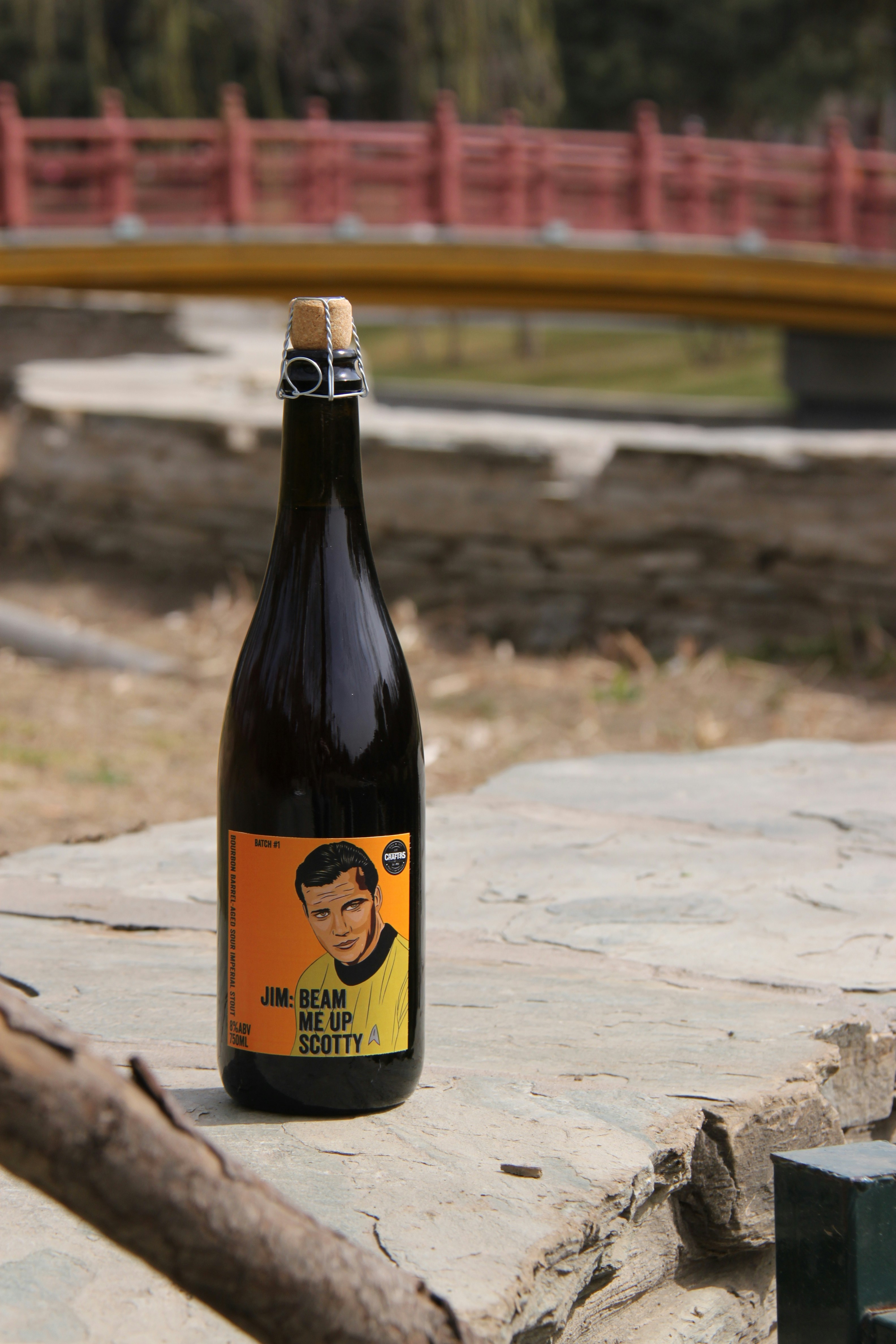 Bottle of Jim Beam whiskey featuring a playful label with a cartoon character and a clever tagline, placed on a stone surface with a blurred bridge in the background.