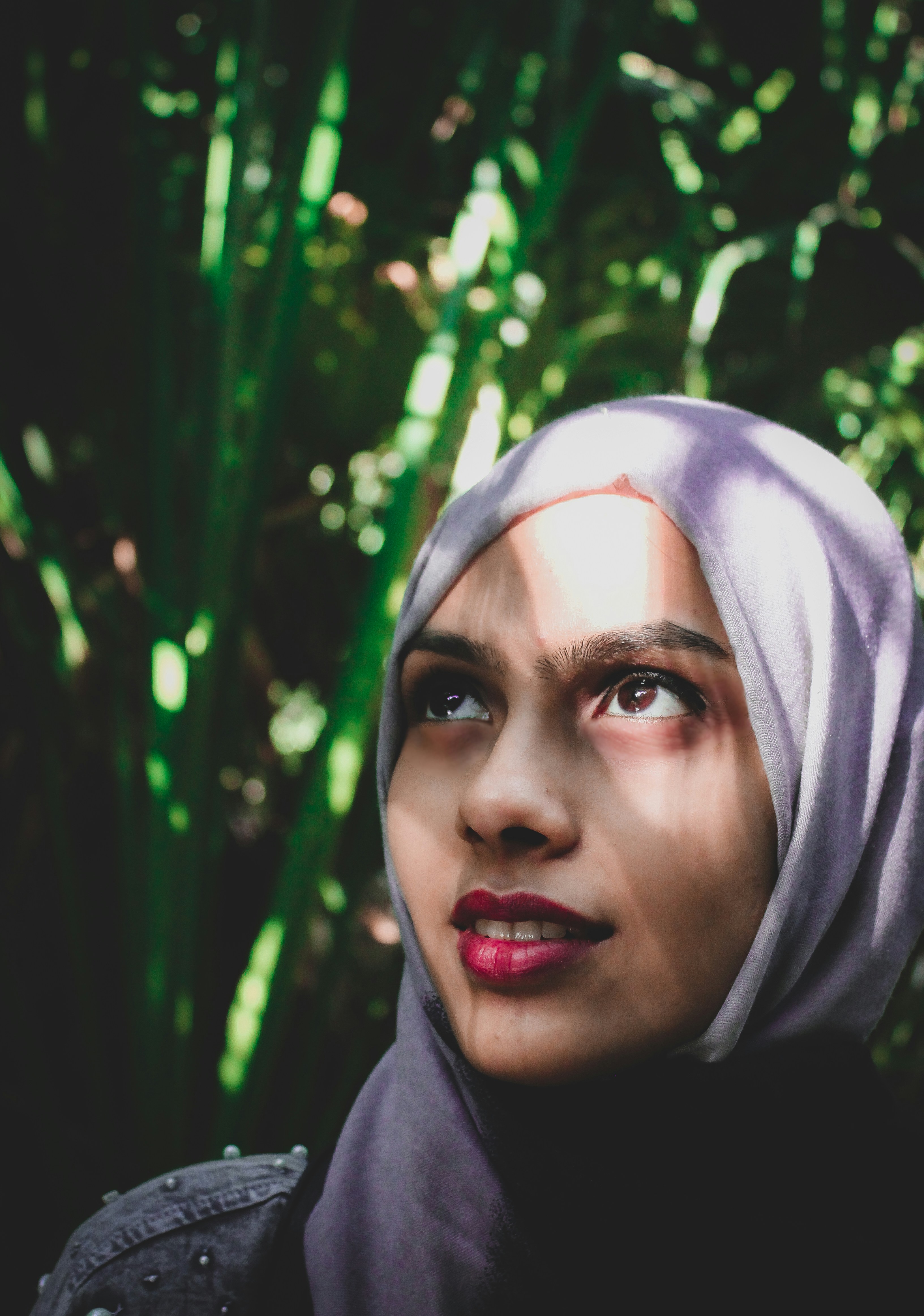 A young woman gazes upward, her face illuminated by dappled sunlight filtering through lush greenery. The serene expression contrasts with the vibrant backdrop.