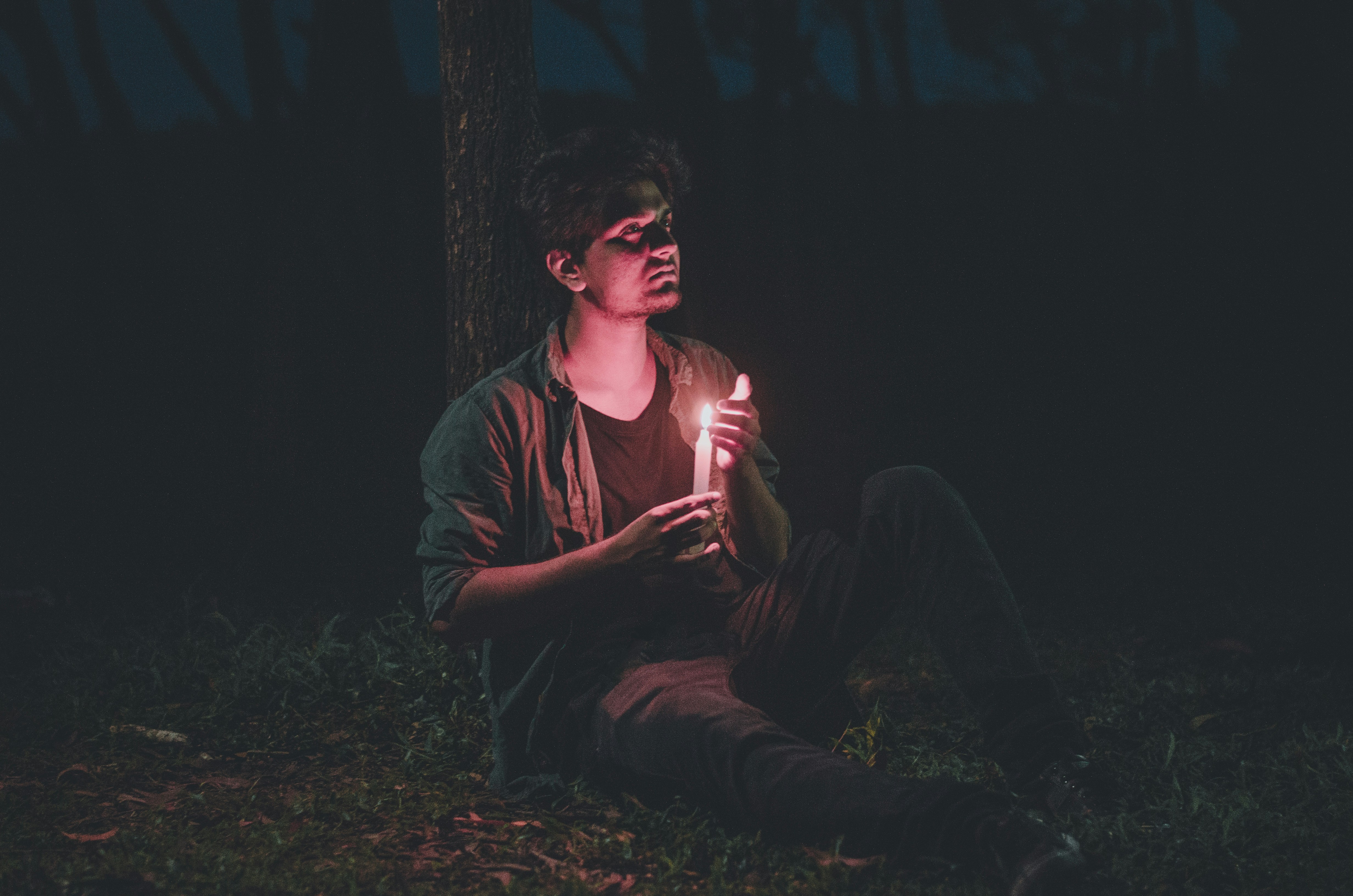 Person sitting against a tree in the dark, holding a lit candle that casts a warm glow on their face.