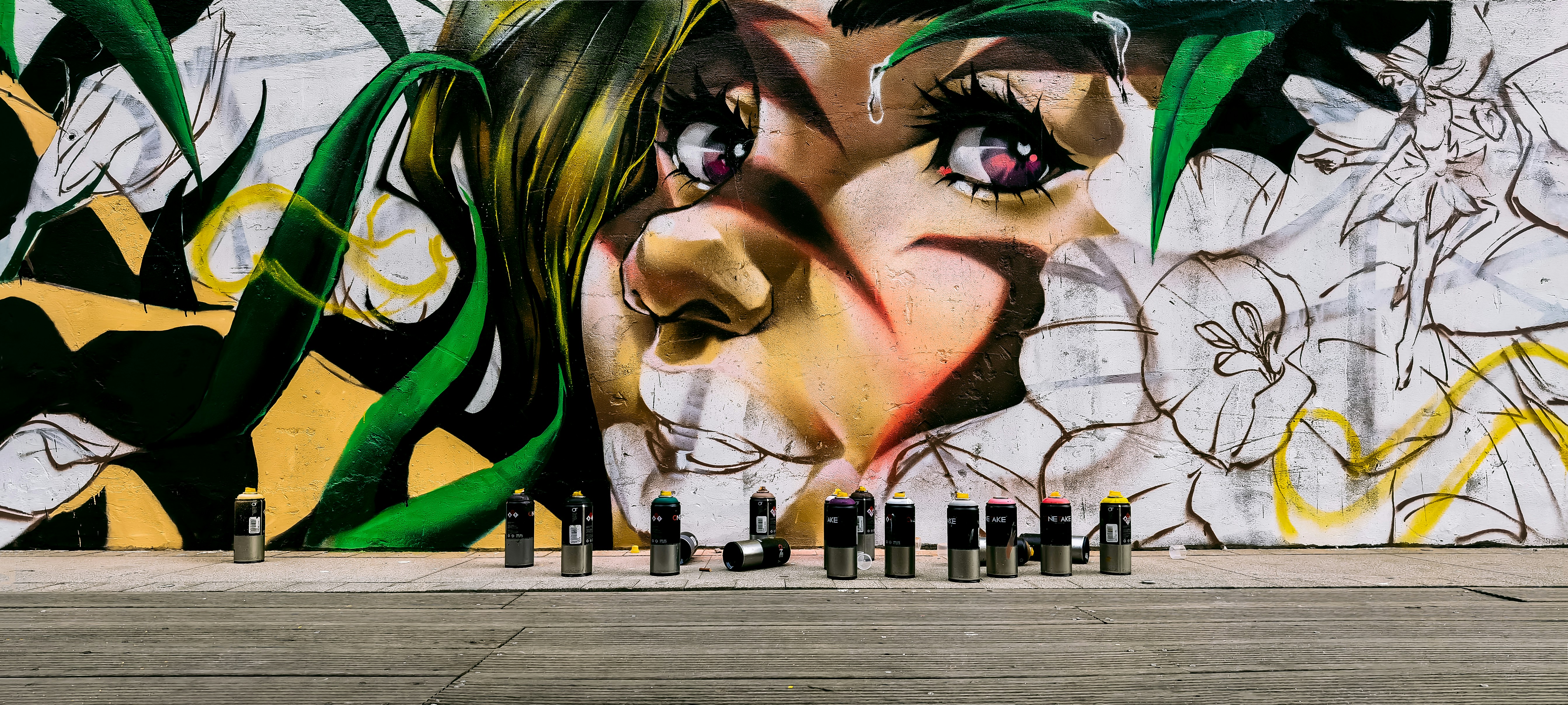Vibrant graffiti mural with a central face motif, accented by spray cans lined up on a wooden platform; the composition emphasizes color and urban texture.