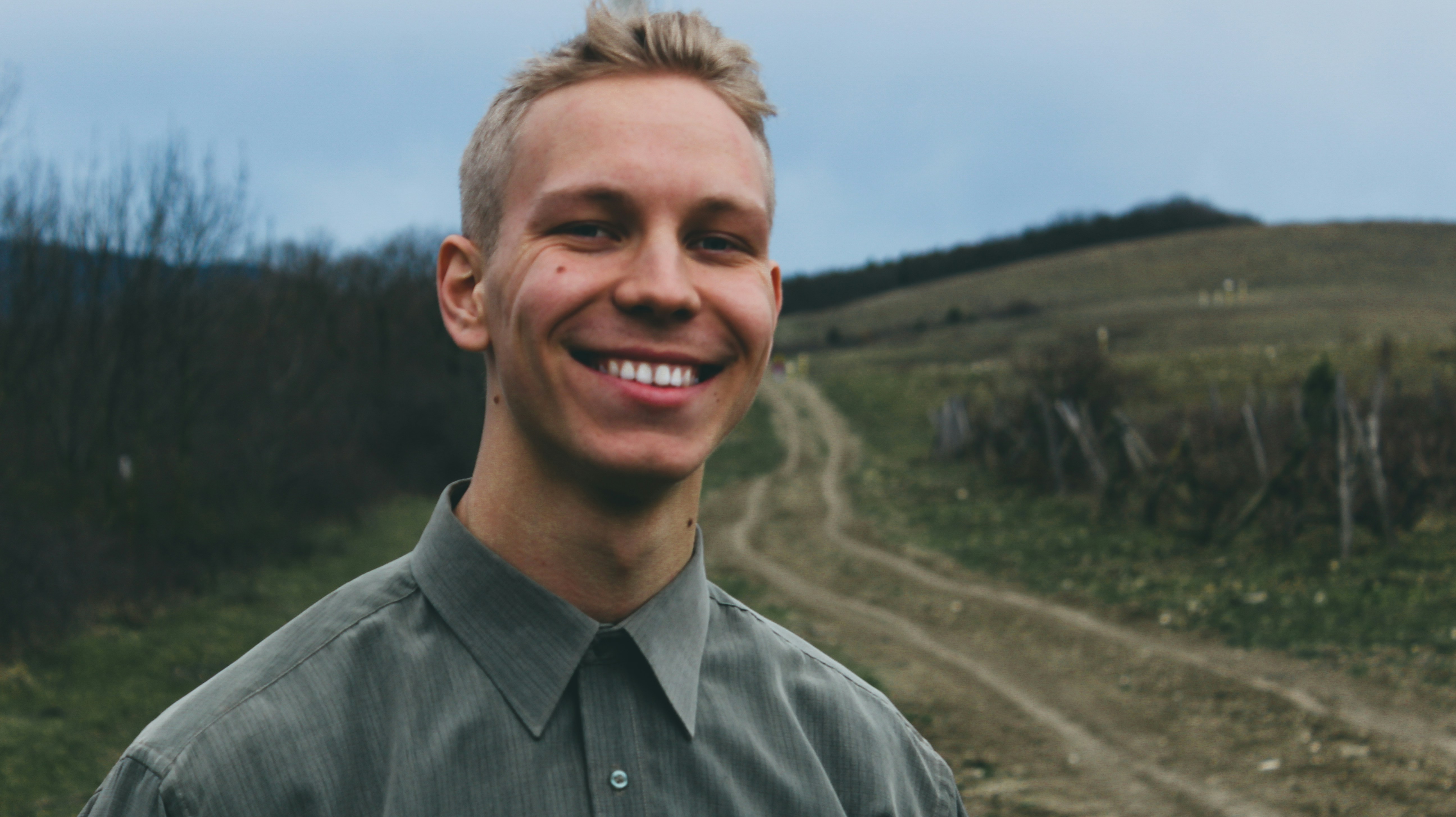 Smiling young man stands confidently on a dirt path surrounded by rolling hills and trees, capturing a moment of happiness in a serene landscape.