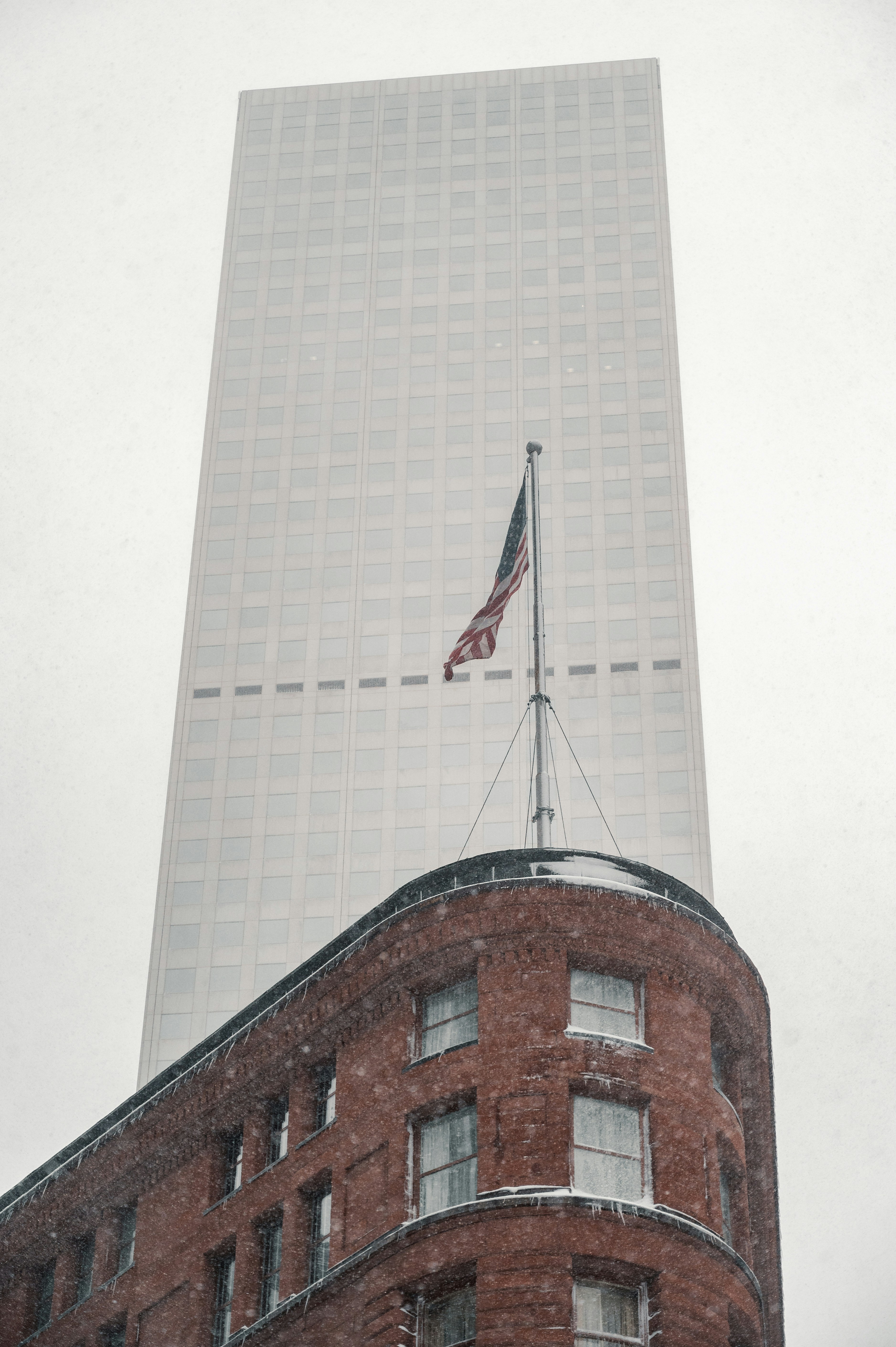 Historic brick building with rounded edges beneath a towering glass skyscraper, featuring an American flag amidst falling snow.