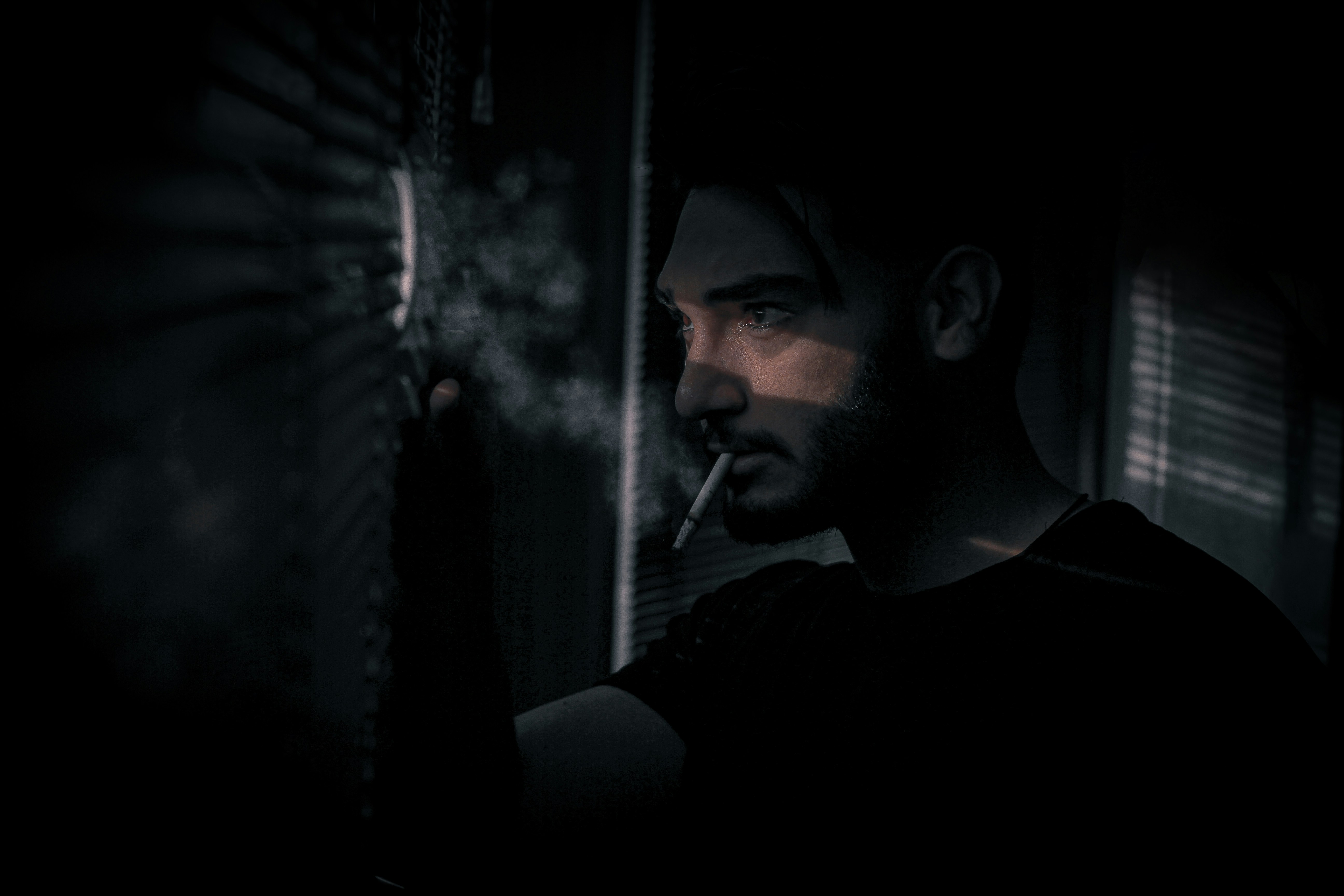 A contemplative figure gazes through blinds, cigarette in hand, as soft light creates a contrast of shadows and highlights.