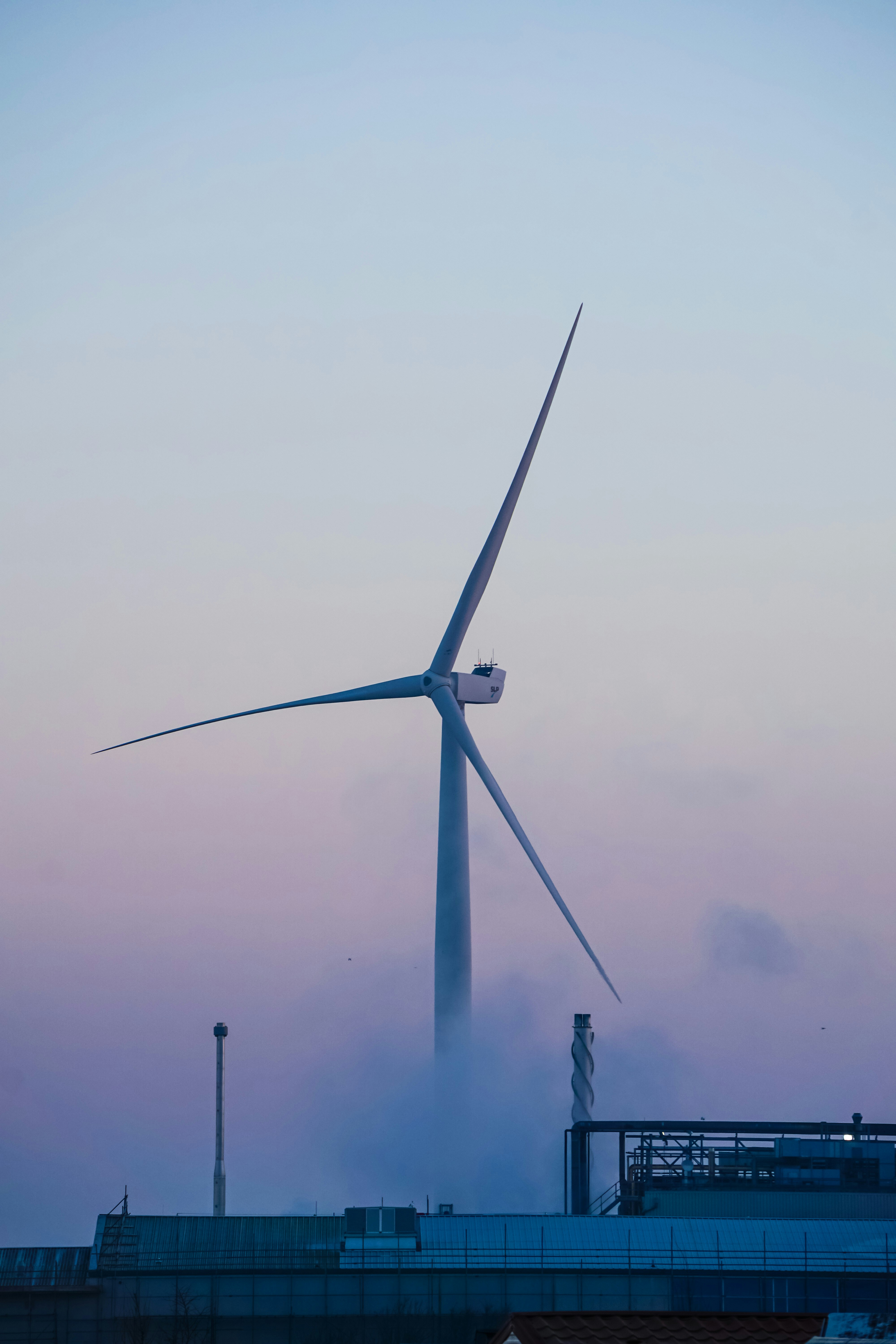 A towering wind turbine stands against a pastel sky, surrounded by industrial structures and wisps of mist, symbolizing the intersection of nature and technology.