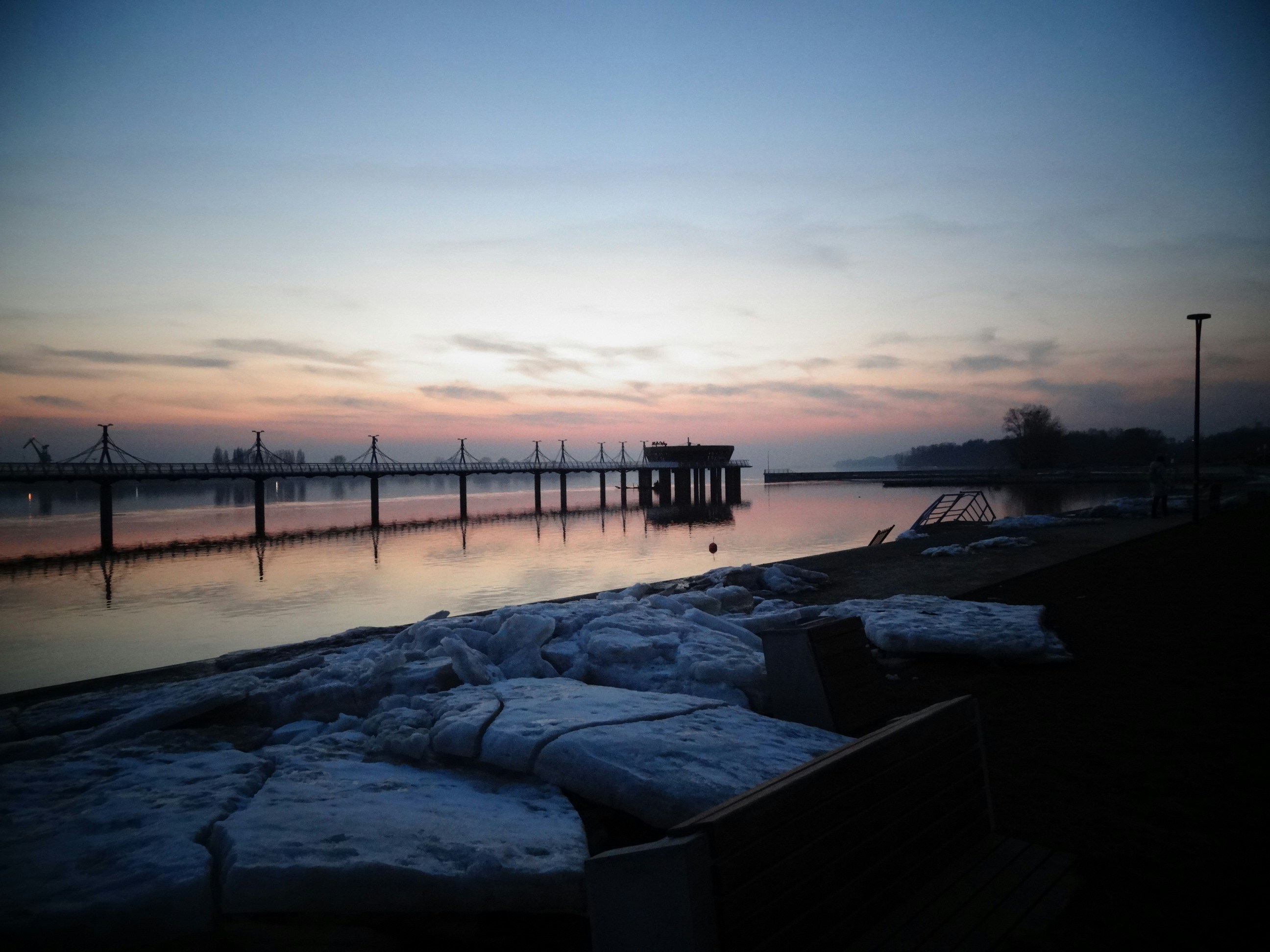 Serene waterfront scene at twilight, featuring a silhouetted pier and soft hues reflecting on the calm water. Ice and snow remnants line the shore.