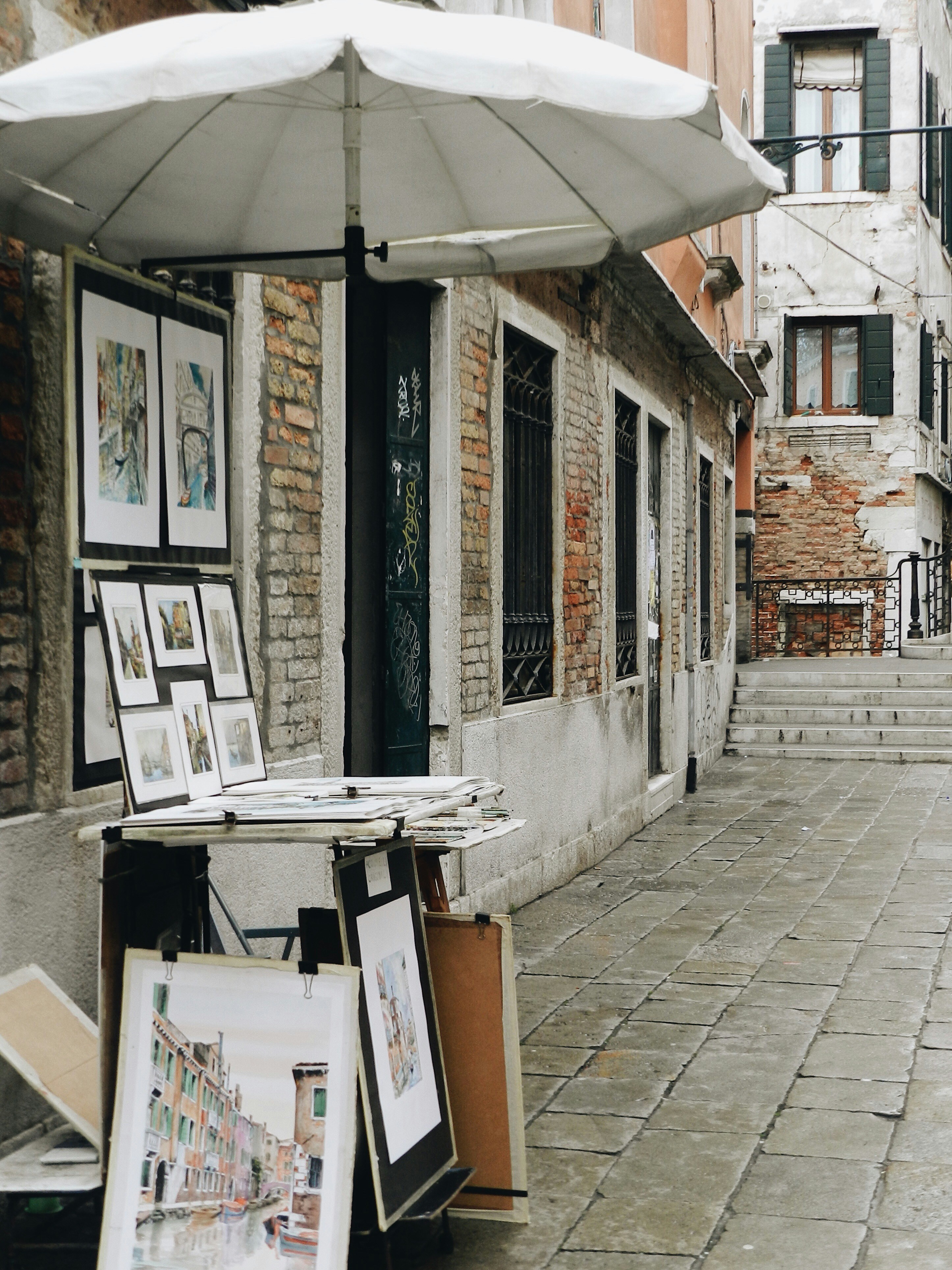 An art stand displaying watercolor paintings in a narrow Venetian alleyway, framed by historic architecture and cobblestone paths.