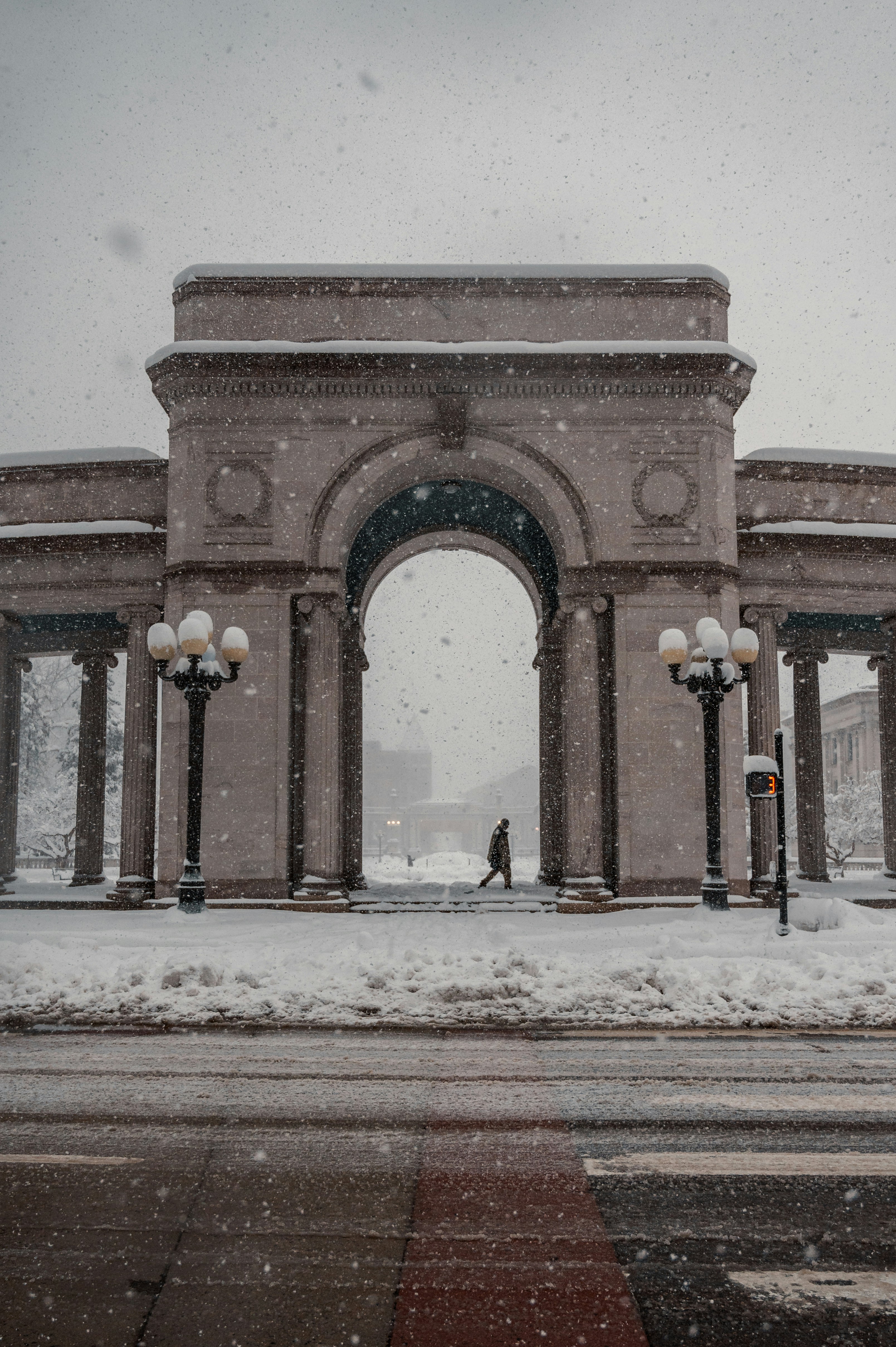A lone figure walks beneath a grand archway during a heavy snowfall, surrounded by a serene winter landscape.