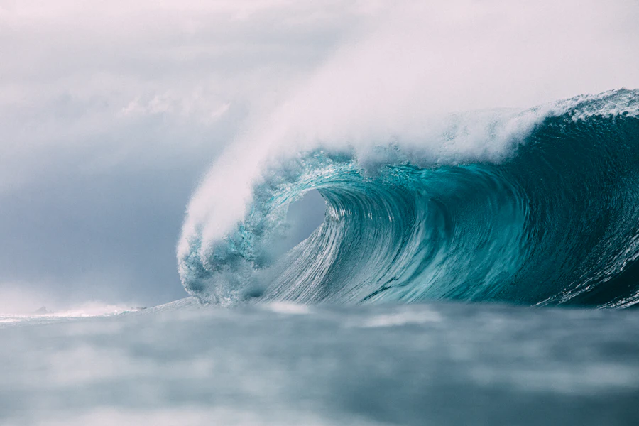 A powerful turquoise wave barrels and begins to curl over, with misty spray and a soft pastel sky behind it.