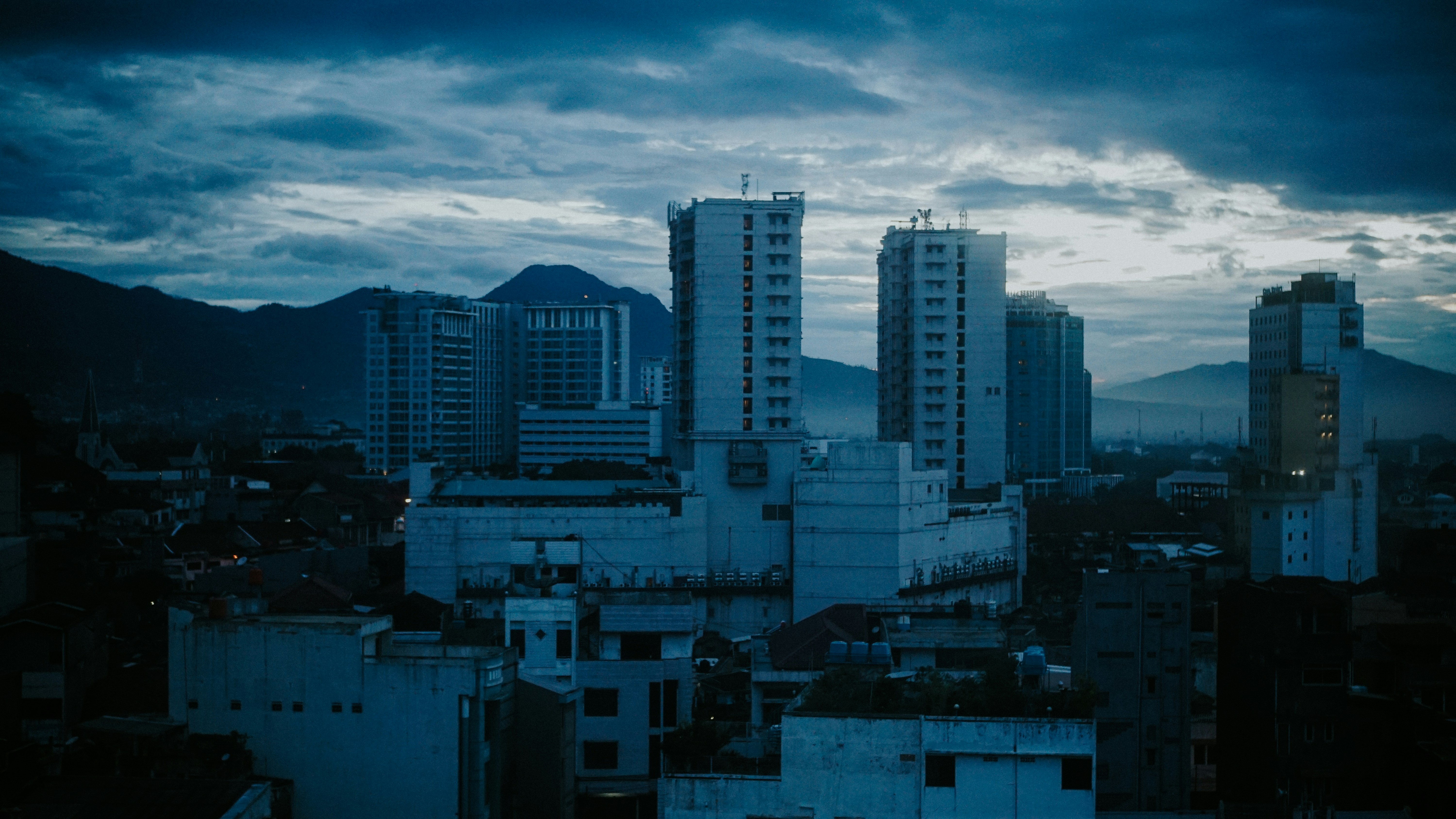 Silhouetted skyline of a bustling city at twilight, framed by distant mountains and a moody sky.