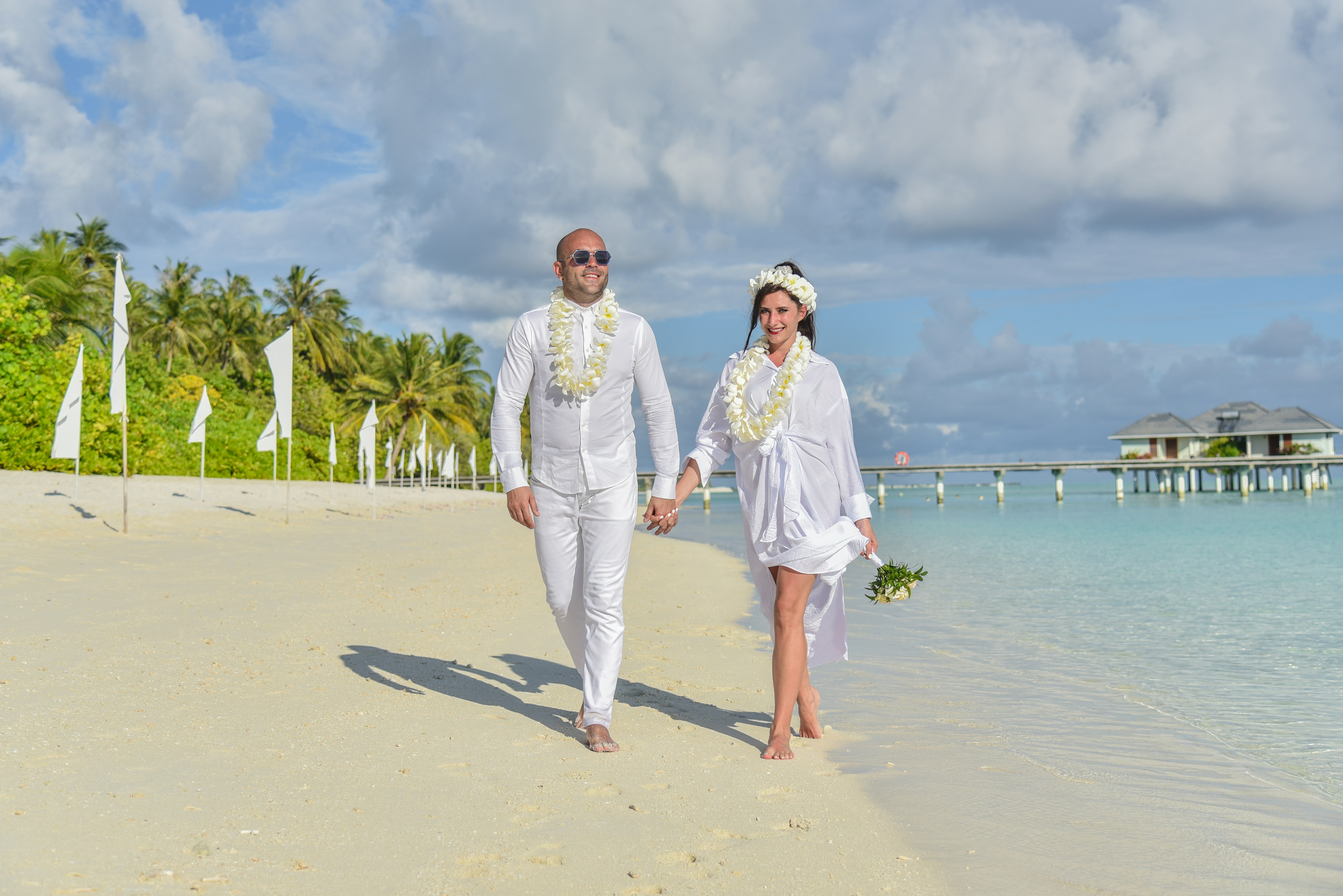 Couple walking hand-in-hand along a pristine beach, adorned in white attire and floral leis, with a backdrop of lush greenery and gentle waves.